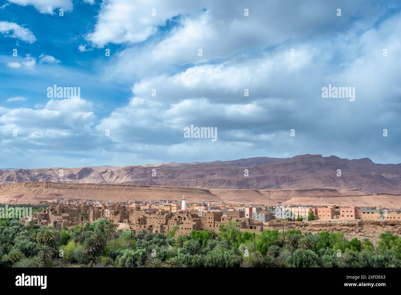 Lush palm trees surround mud-brick buildings against towering cliffs ...
