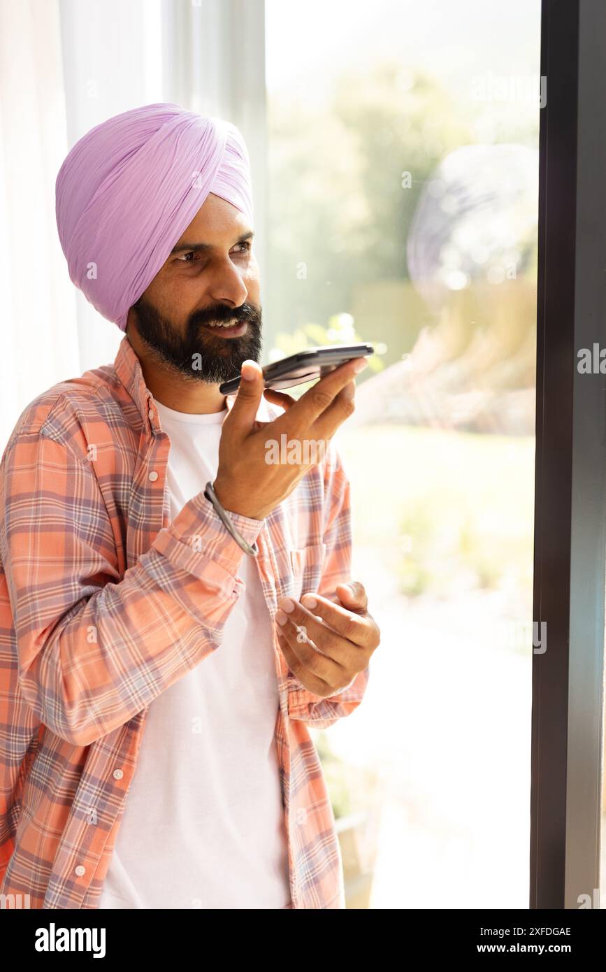 Speaking on smartphone, Indian man in turban standing by window at home ...