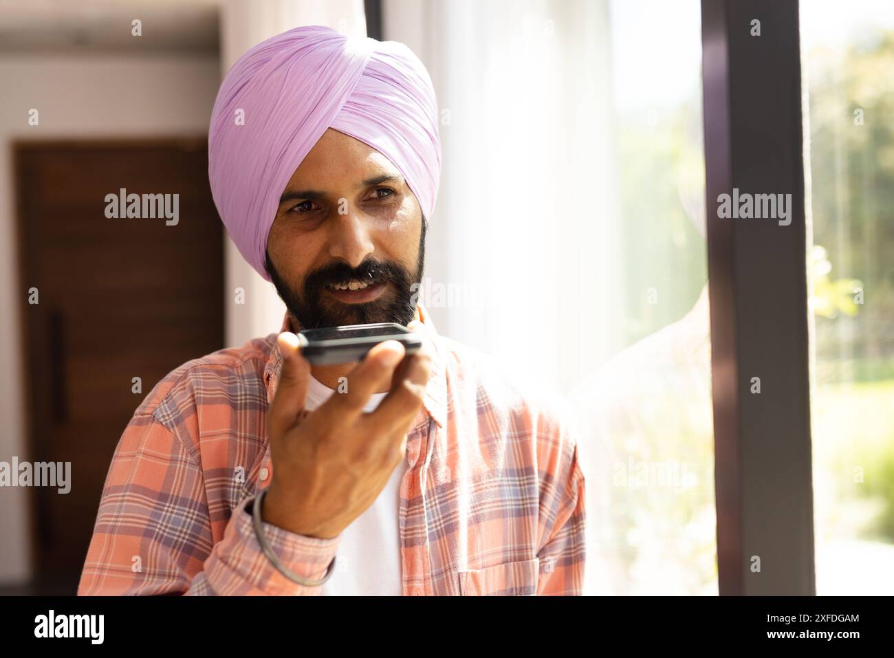 Speaking into smartphone, Indian man in turban standing by window at ...
