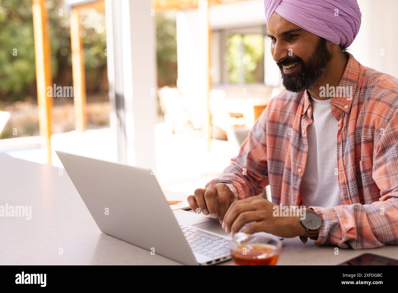 Typing on laptop, smiling man in turban working from home office, copy ...