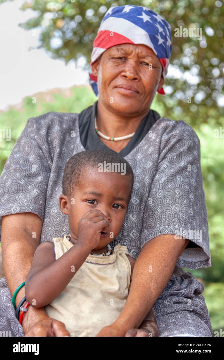old basarwa woman sitting on the sand , child on her lap, in the bush ...
