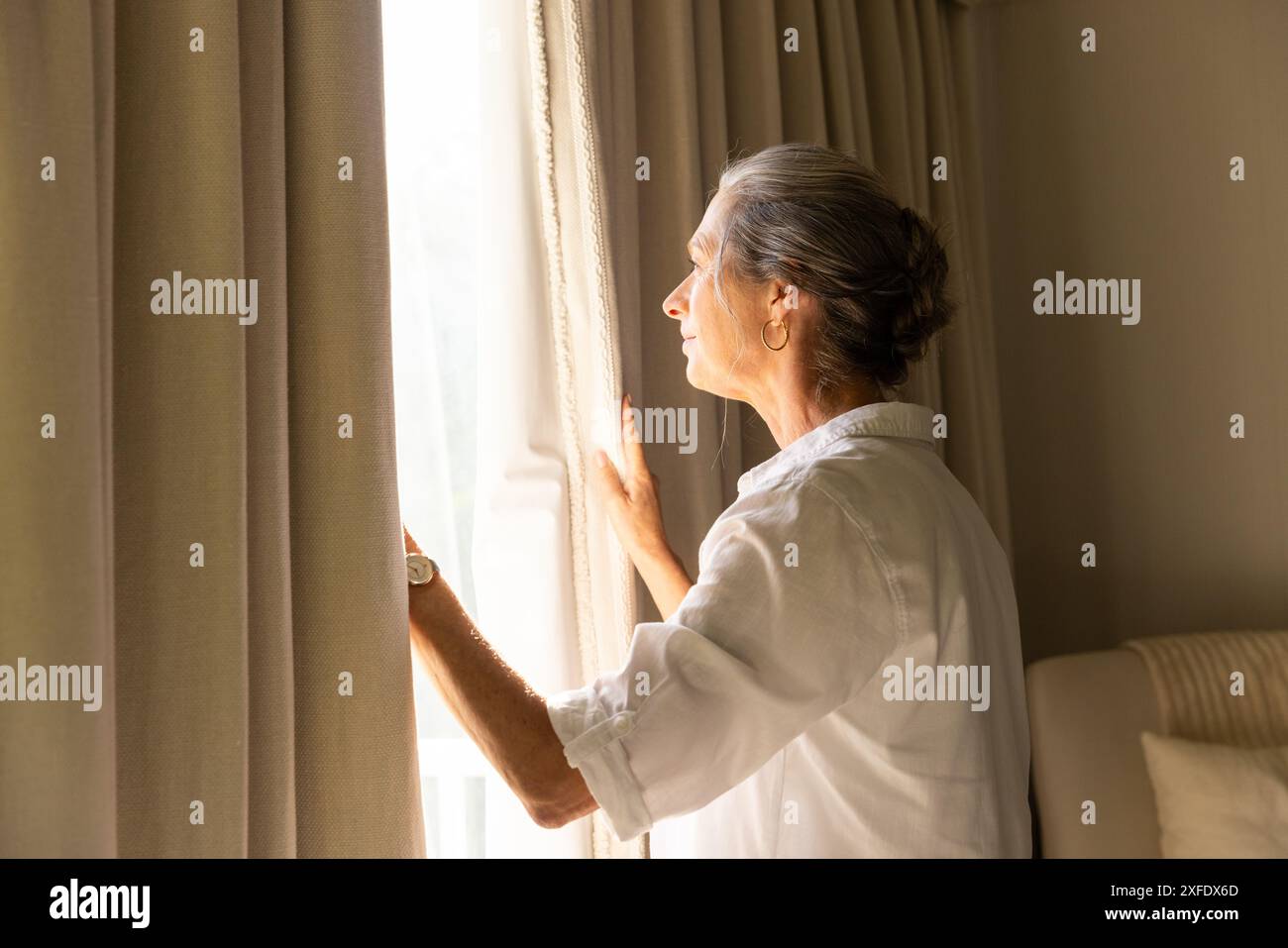 Looking out window, mature woman in white shirt enjoying sunlight at ...