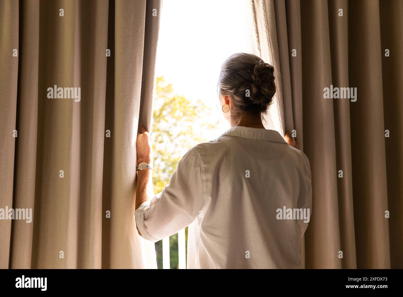 Opening curtains, mature woman in white shirt looking outside window at ...