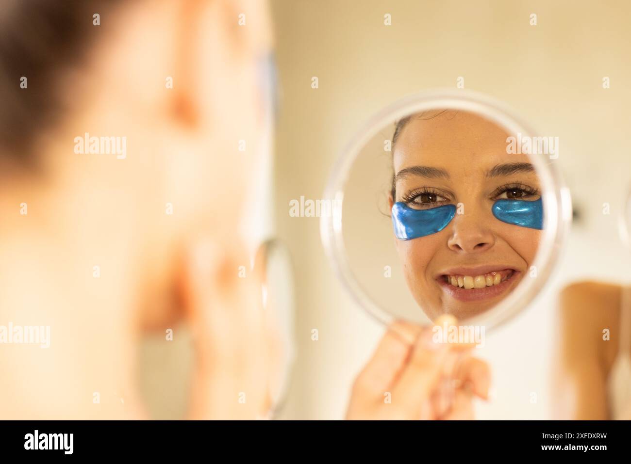 Applying under-eye patches, woman smiling and looking in mirror at home ...