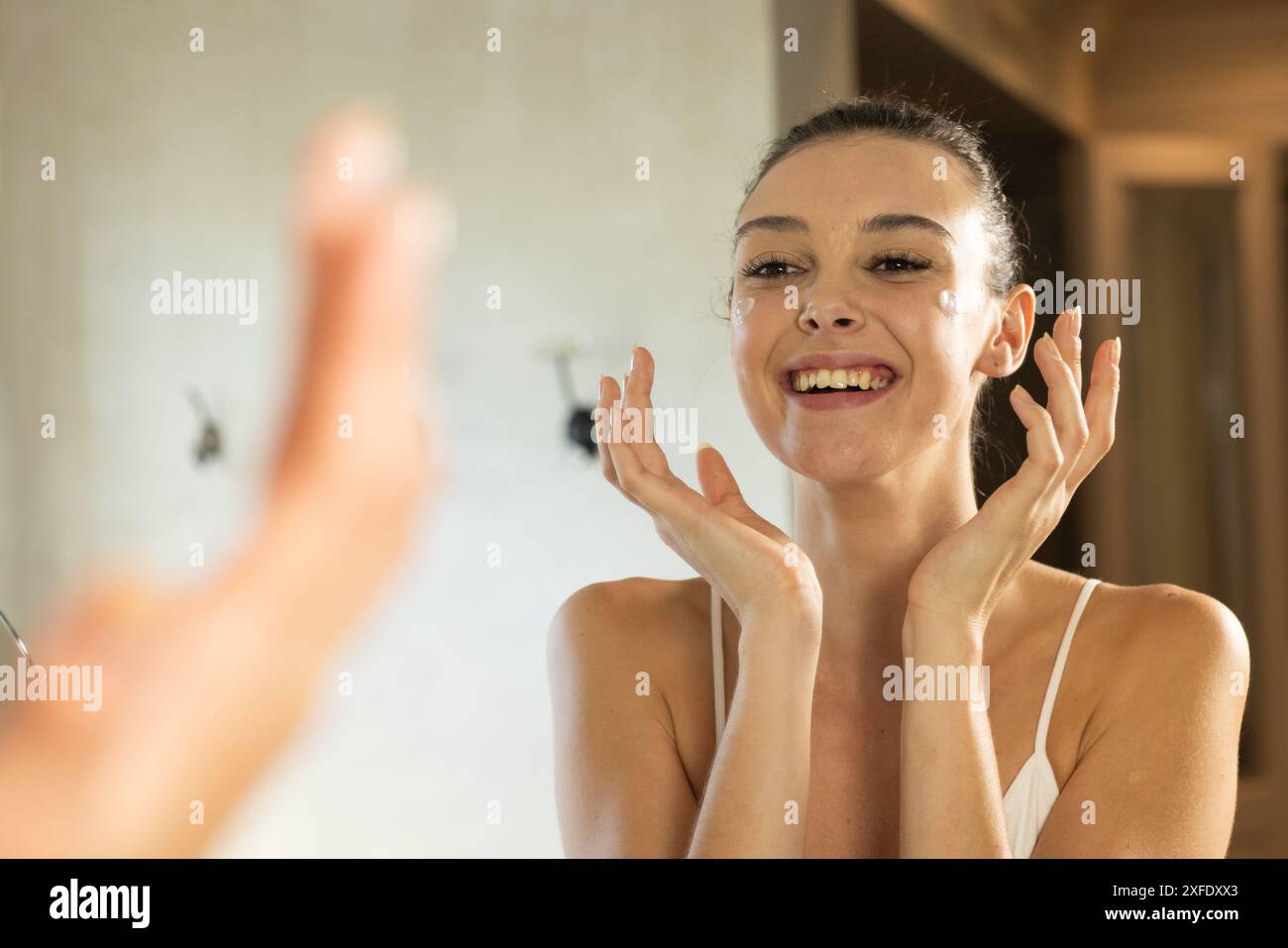 Applying face cream, young woman smiling and looking in bathroom mirror ...