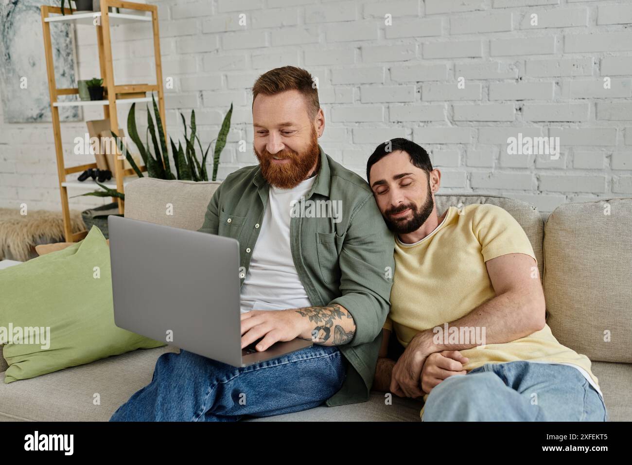 Two men with beards sitting on a couch in a living room, focused on a laptop screen in front of ...