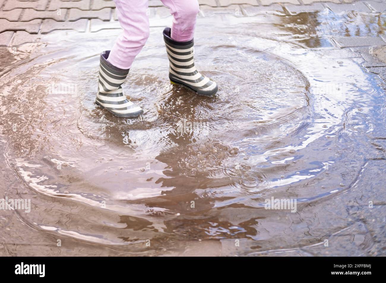 closeup 5-year-old girl joyfully jumps in puddle wearing rubber boots ...