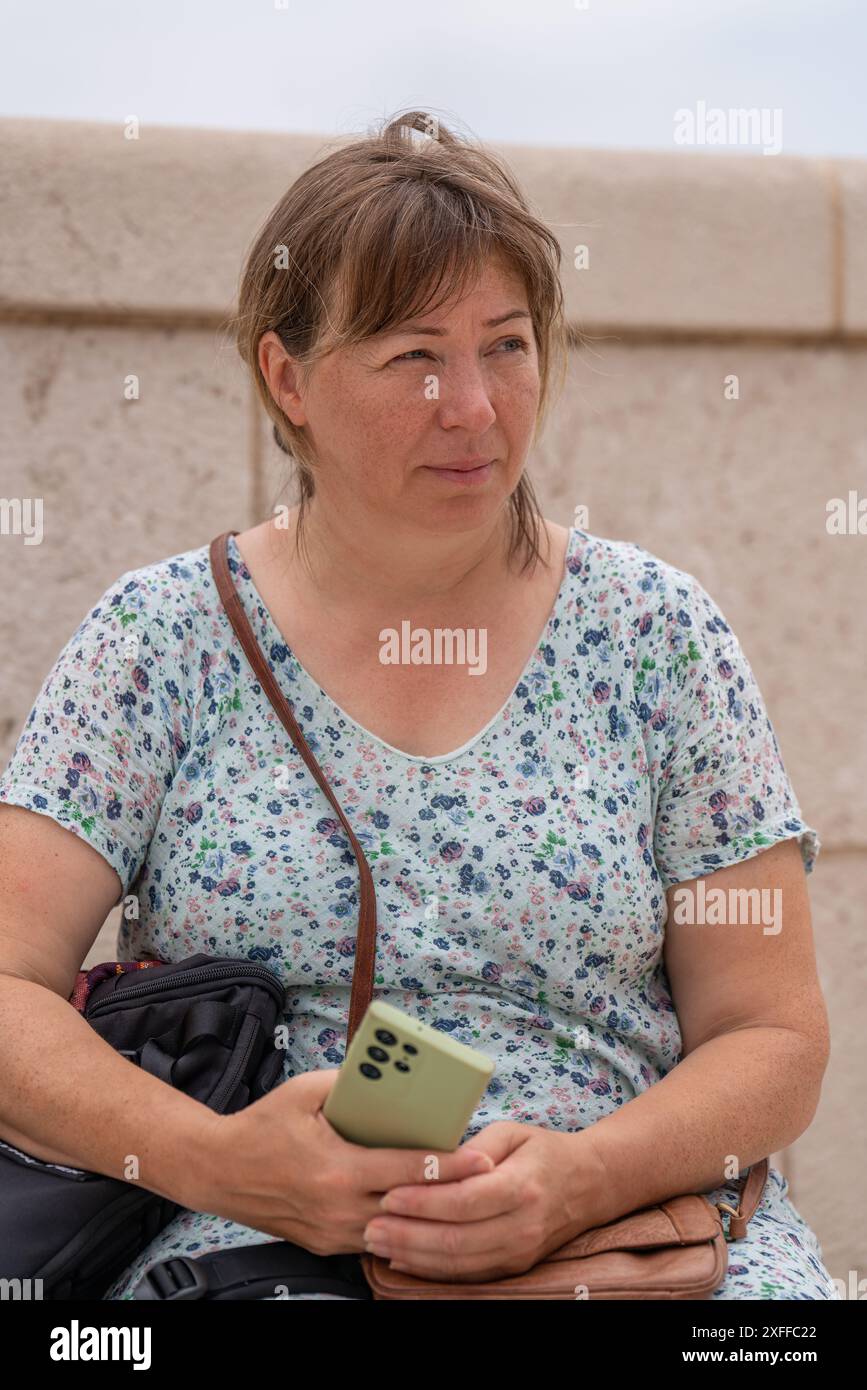 a middle-aged woman holds a mobile phone, sits on the embankment and ...