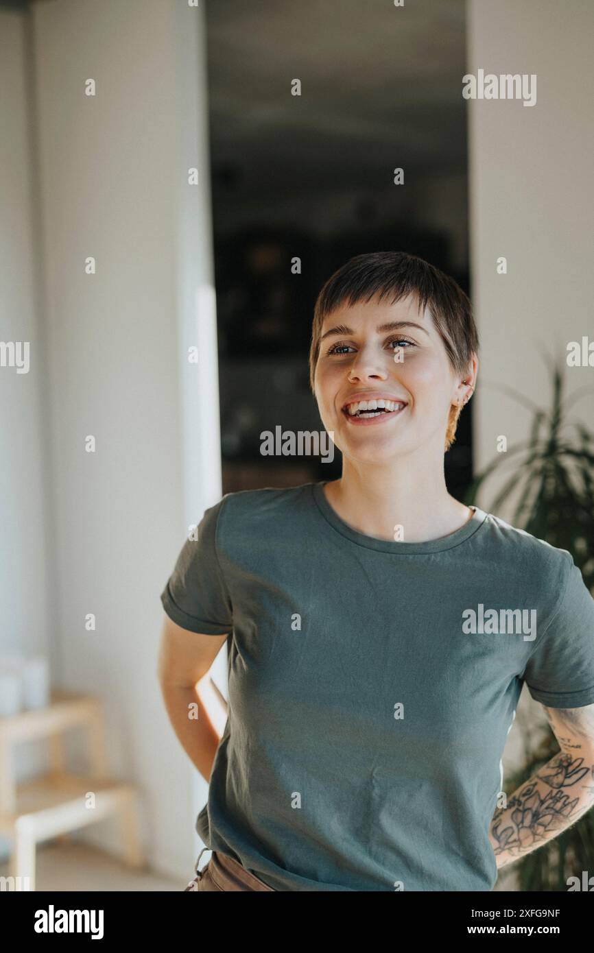 Happy woman with tattooed arm standing with hands behind back at home ...