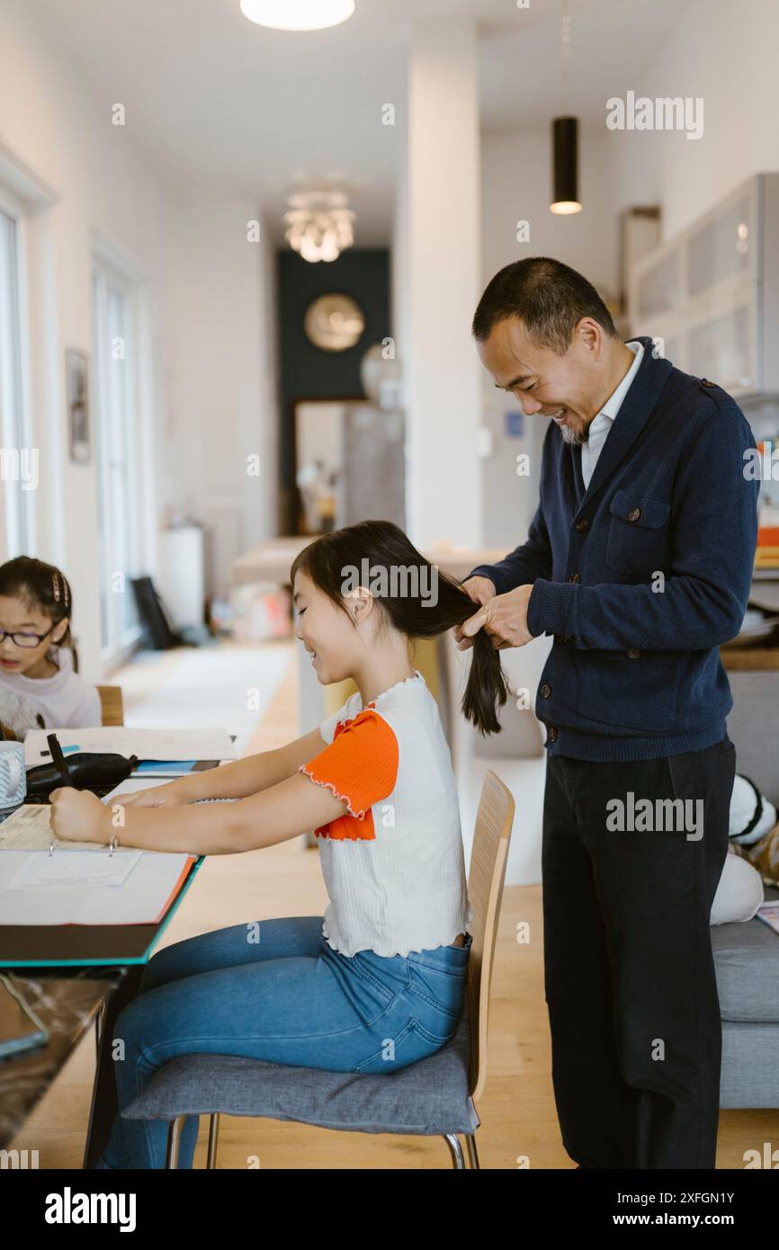 Smiling man tying hair of daughter sitting on chair and doing homework ...
