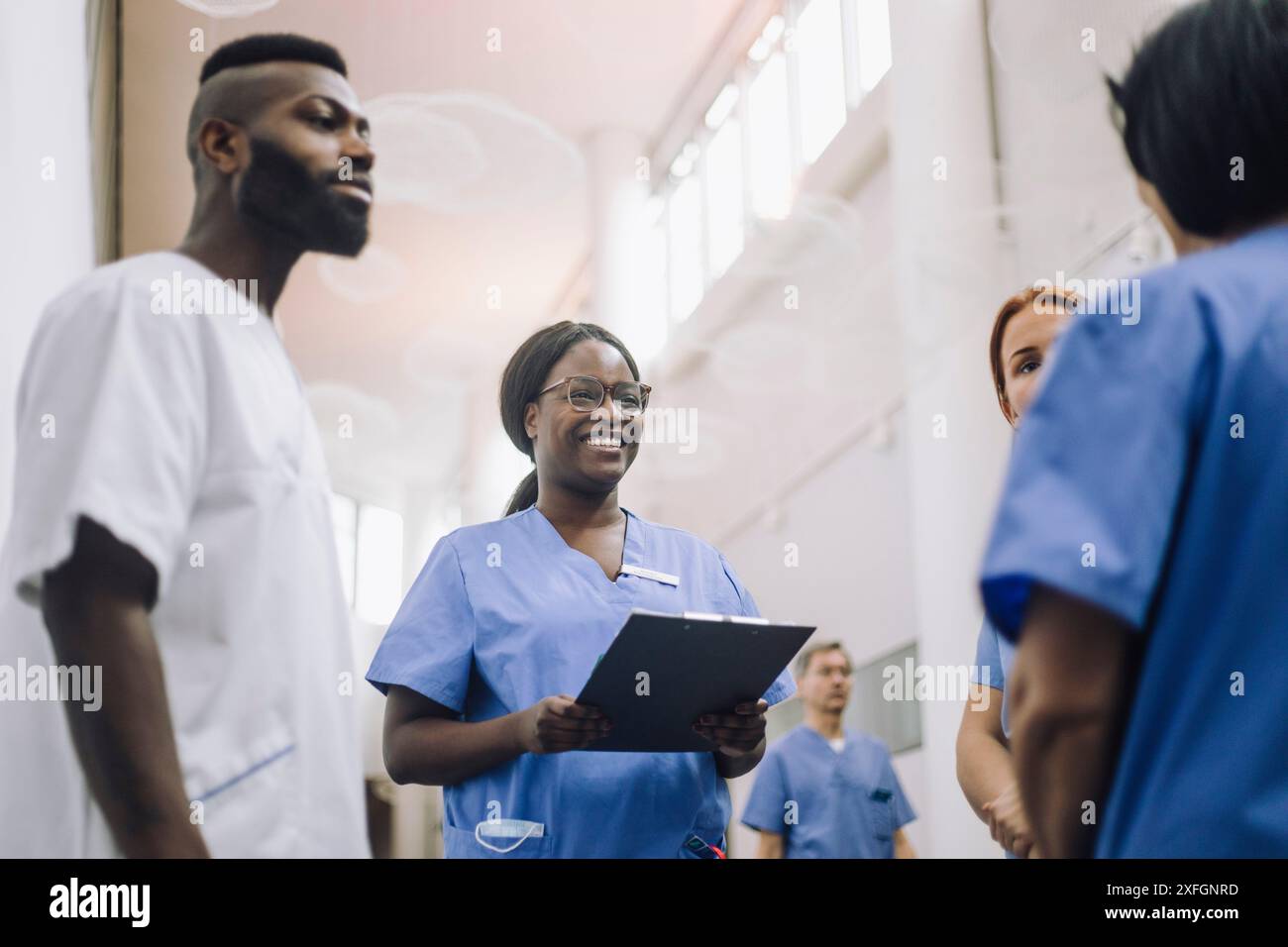 Smiling female nurse holding clipboard while talking with colleagues in ...