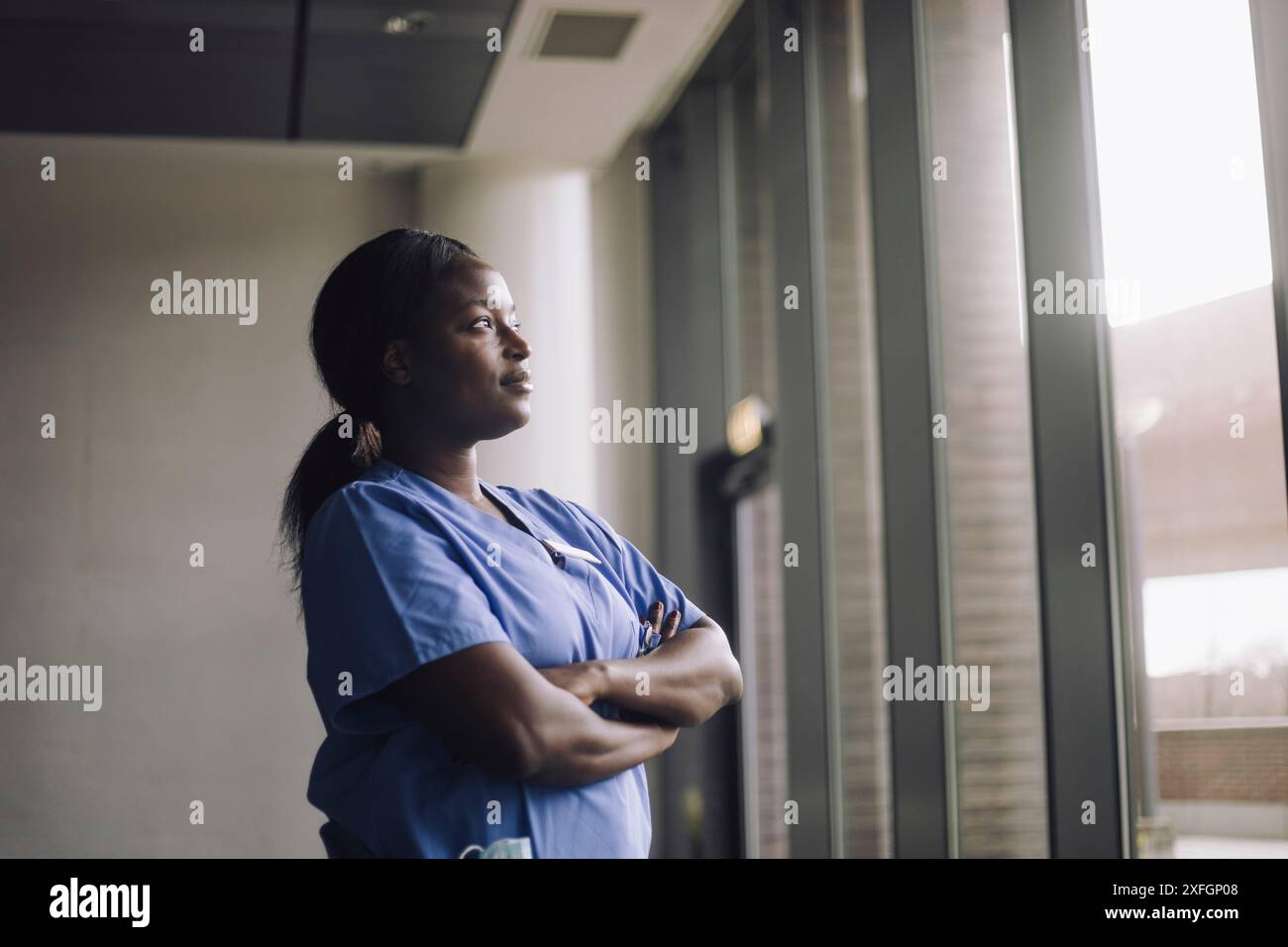 Female medical expert looking through window while standing with arms crossed in hospital Stock ...