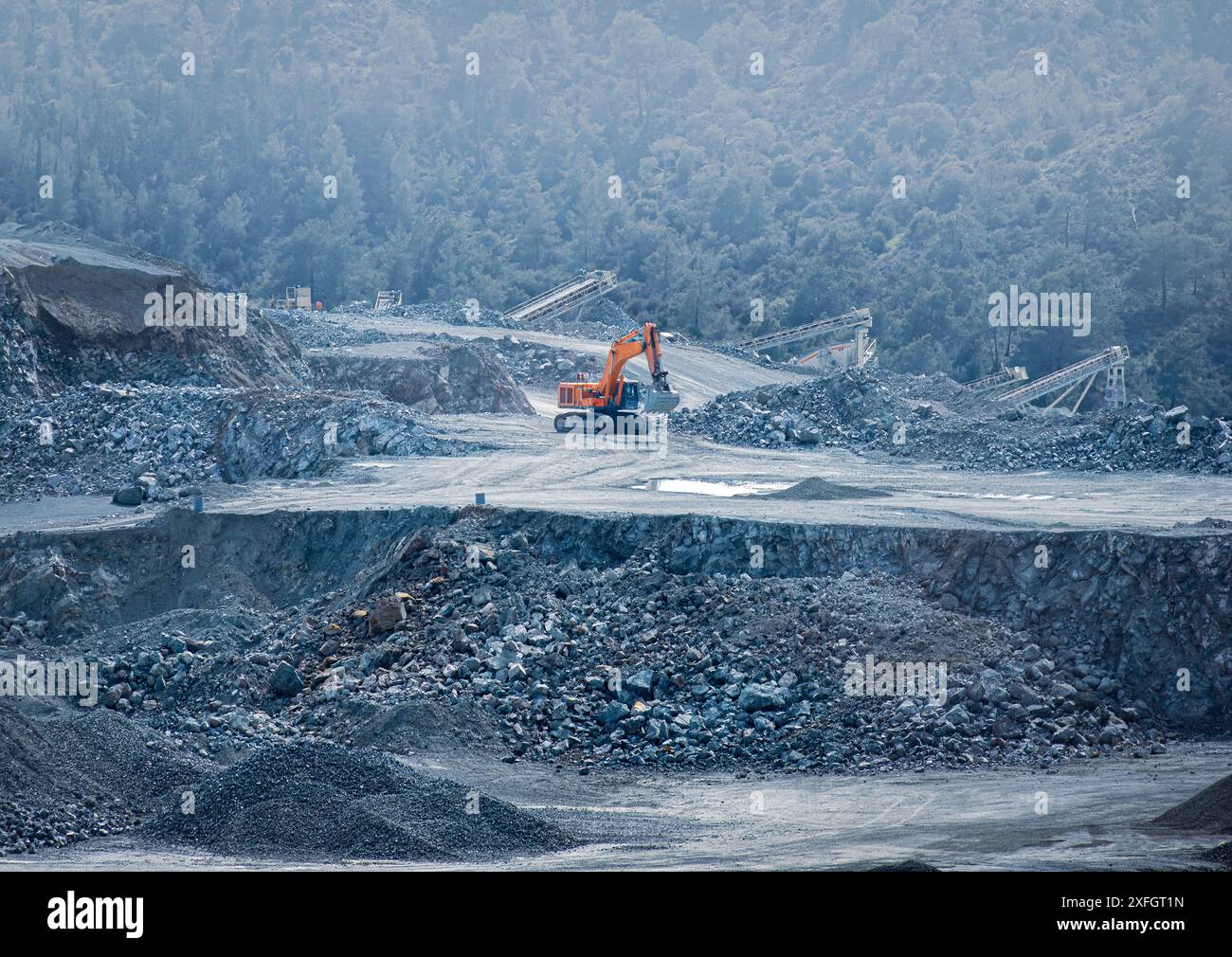 Excavator operating in a diabase quarry with conveyor belts and forest ...