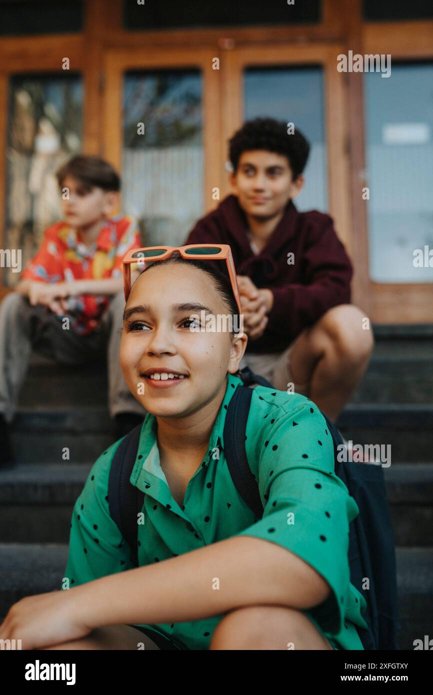 Smiling girl wearing backpack sitting with male friends on staircase in ...