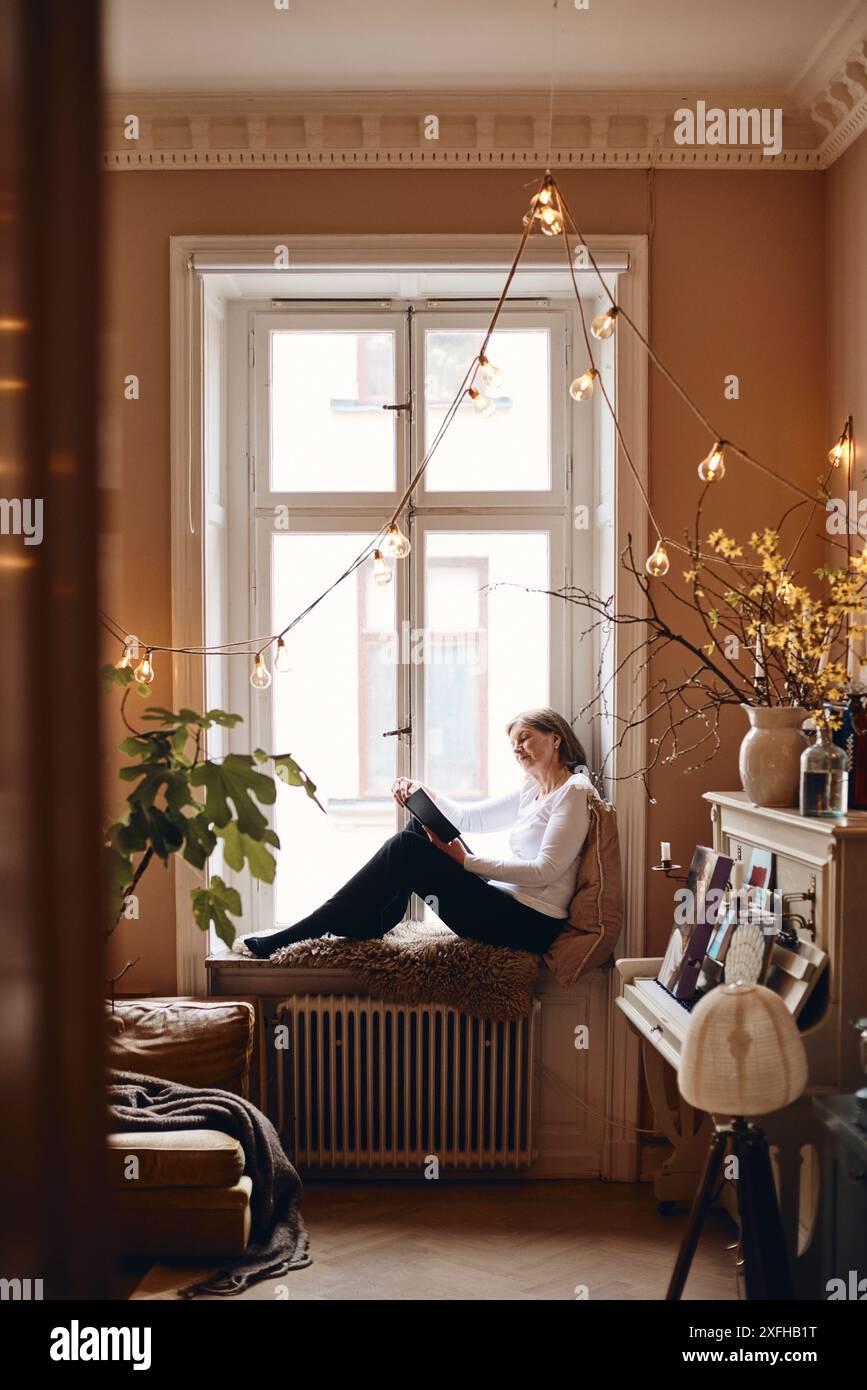 Side view of elderly woman reading book while sitting on window sill ...