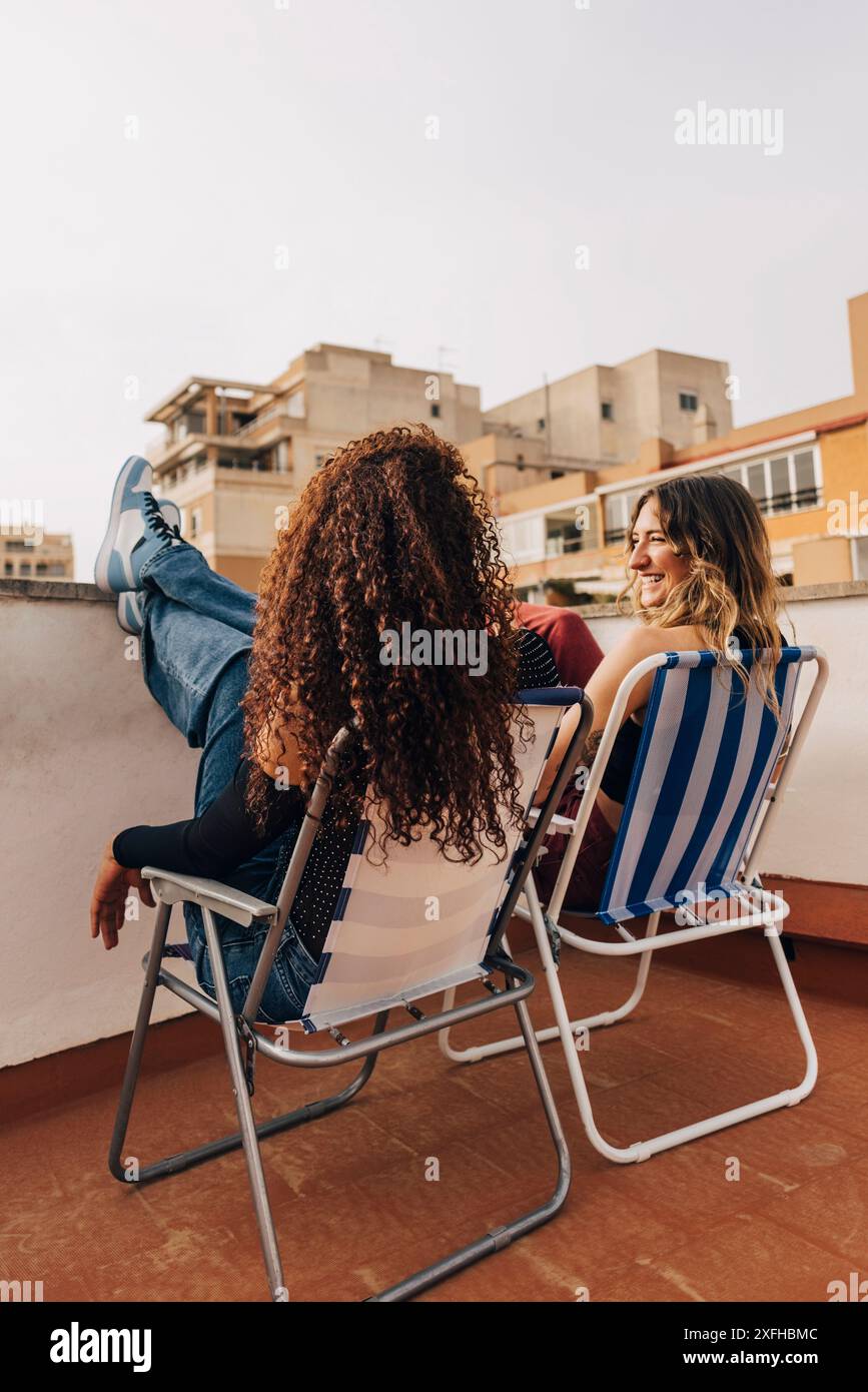 Smiling young woman sitting on chair next to female friend on rooftop ...