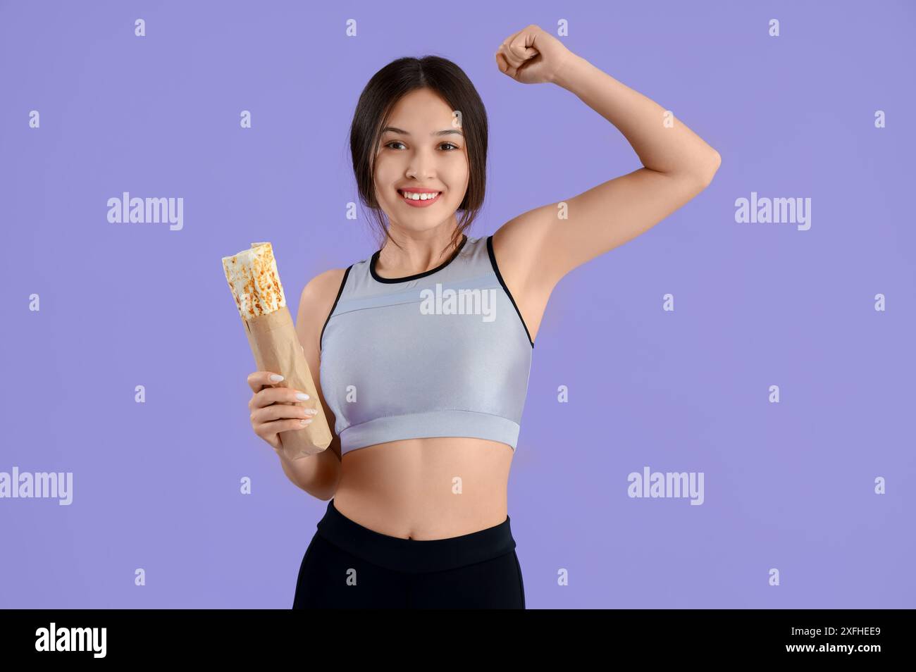 Sporty young Asian woman with doner kebab showing muscles on lilac ...