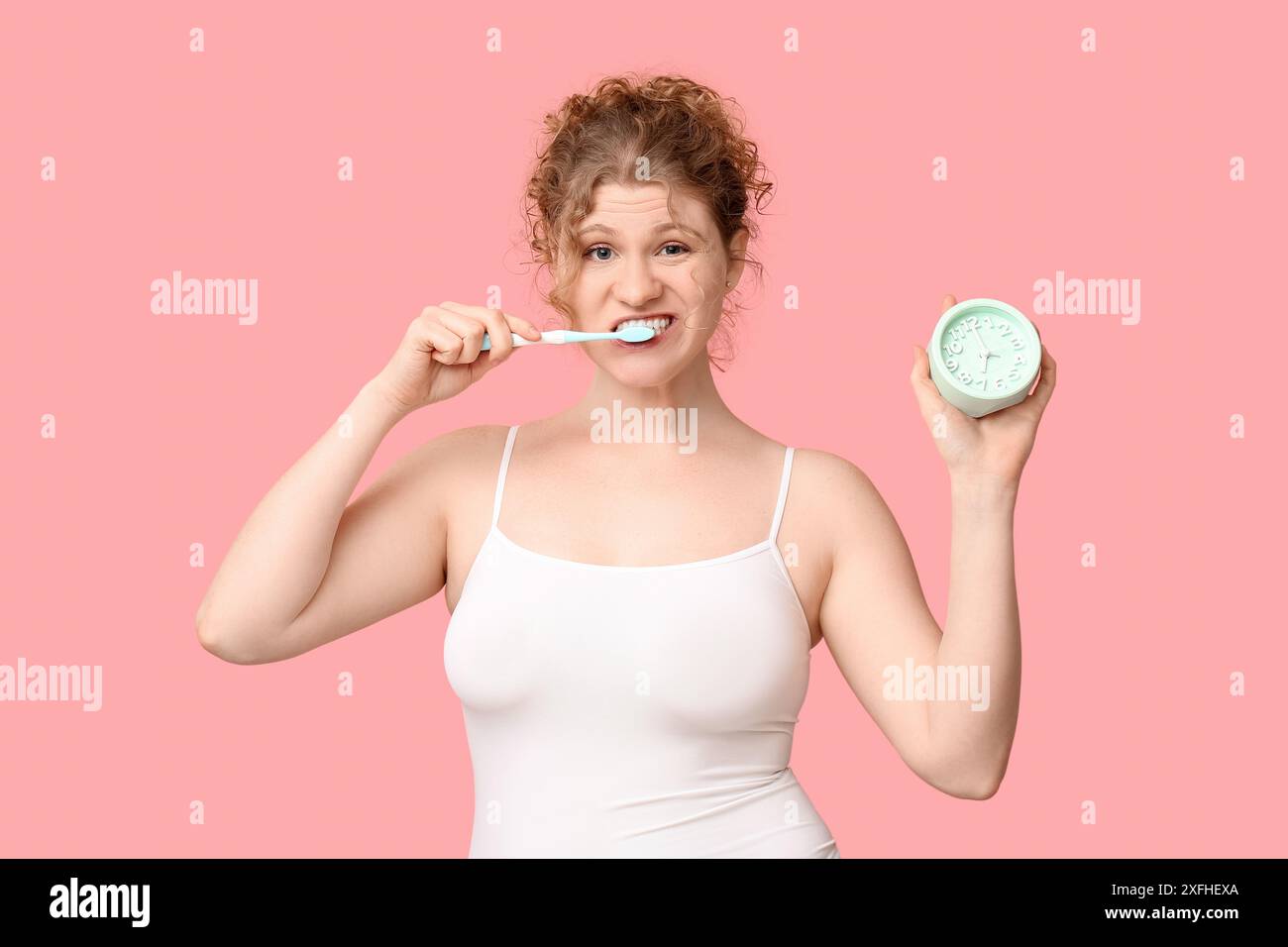 Beautiful young woman with alarm clock brushing teeth on pink ...