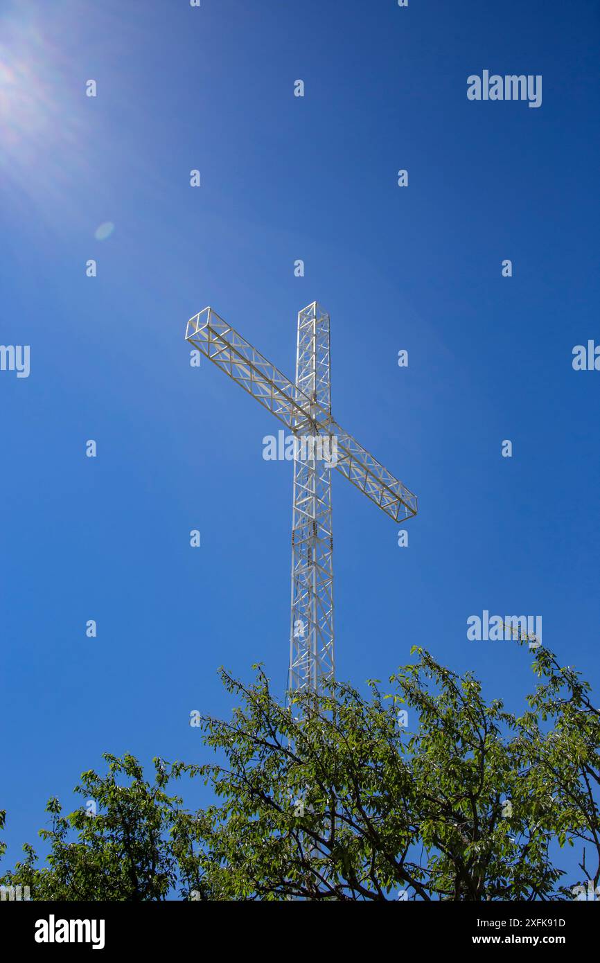 High metal cross structure over tree tops, with blue sky and sun rays ...