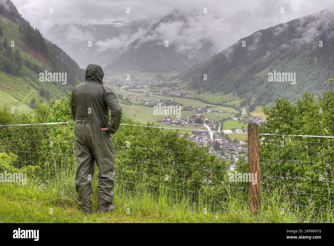 In the Austrian Alps, a hiker in a rain suit stands at a viewpoint ...