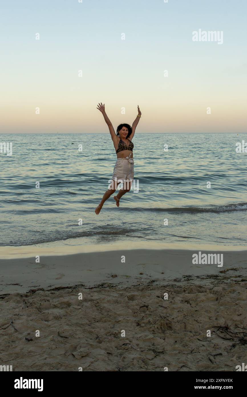 This image captures a woman mid-jump on a sandy beach. Her joyful pose ...