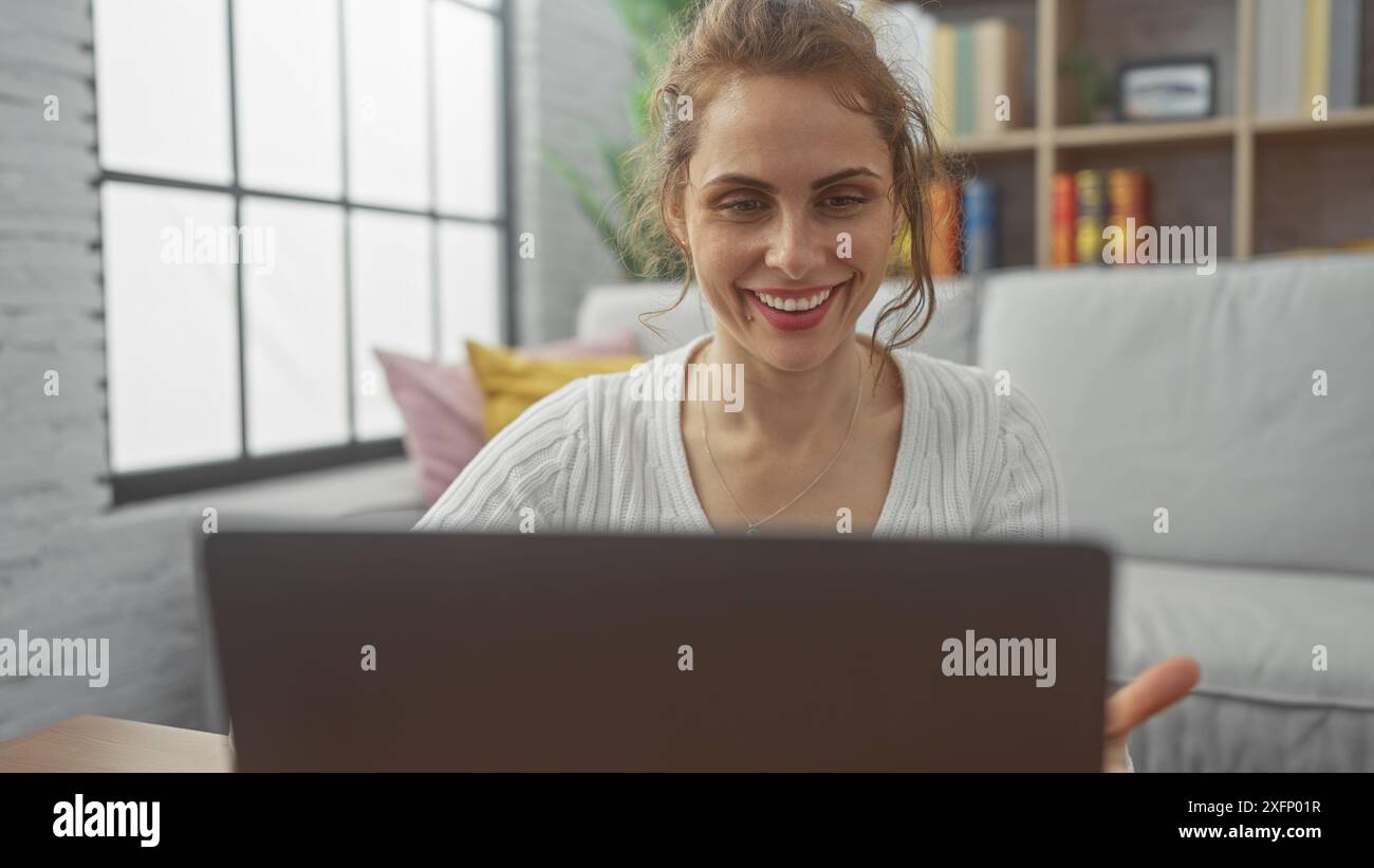 A caucasian woman smiles while working on her laptop in a cozy, sunlit ...