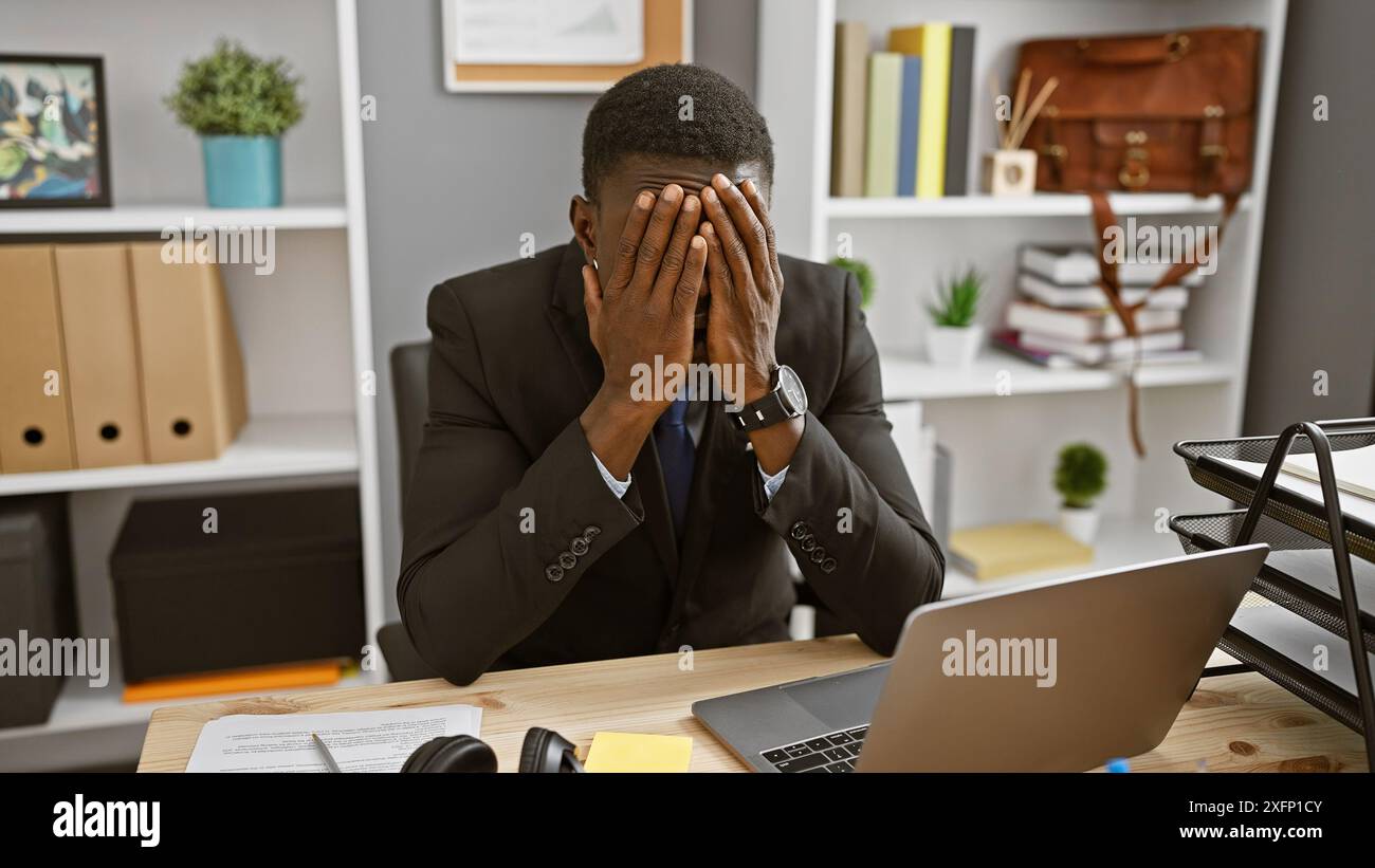 Stressed african man sitting in office with hands on face, laptop and ...