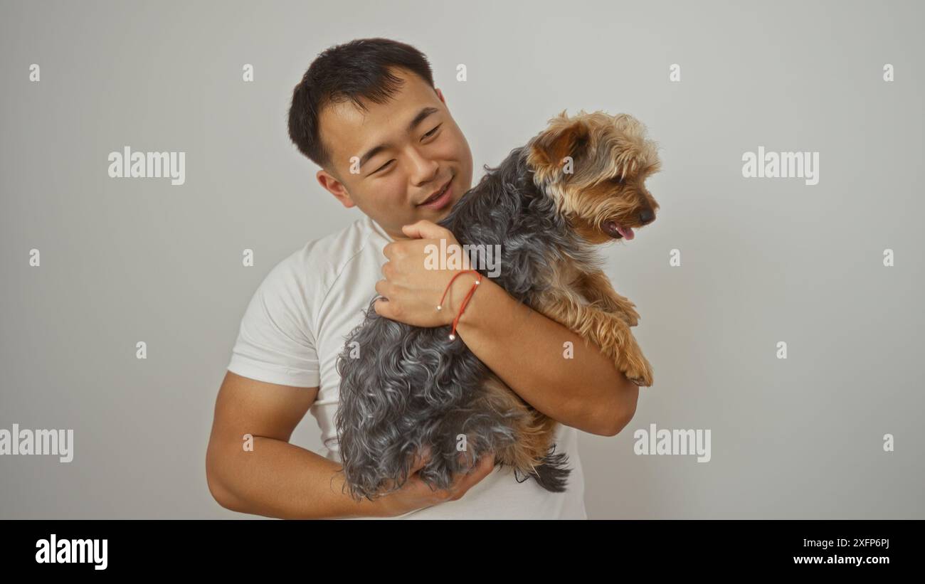 A young chinese man joyfully holds his pet dog against a white wall ...