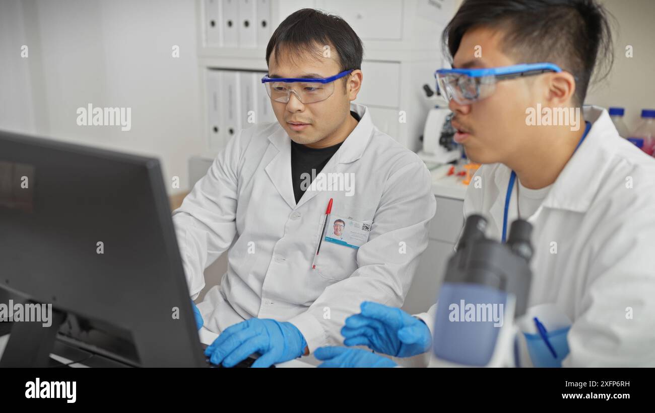 Two men in lab coats and safety glasses working on a computer in a ...