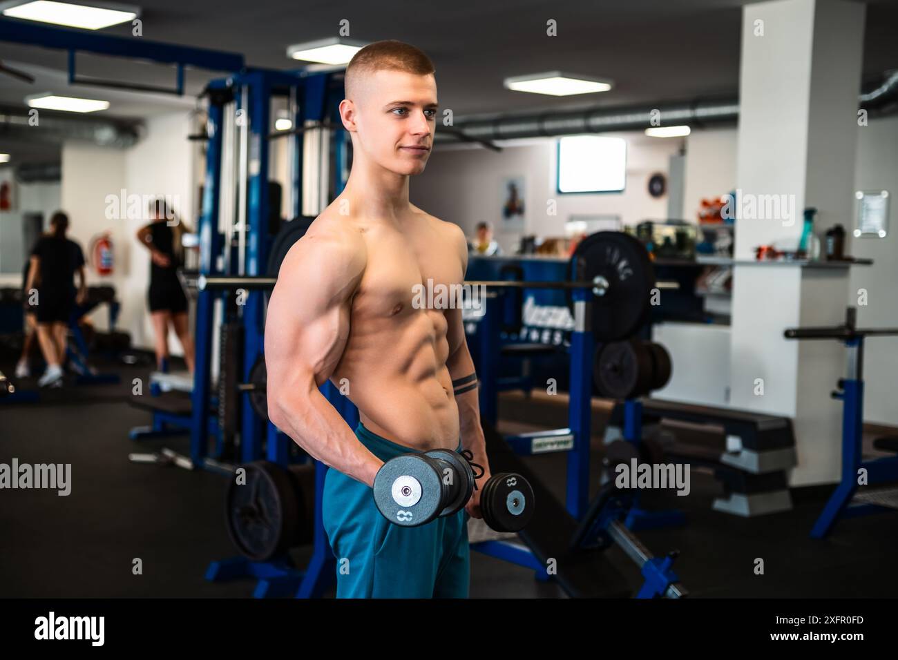 A young blond man in a gym lifts weights with a focused expression ...