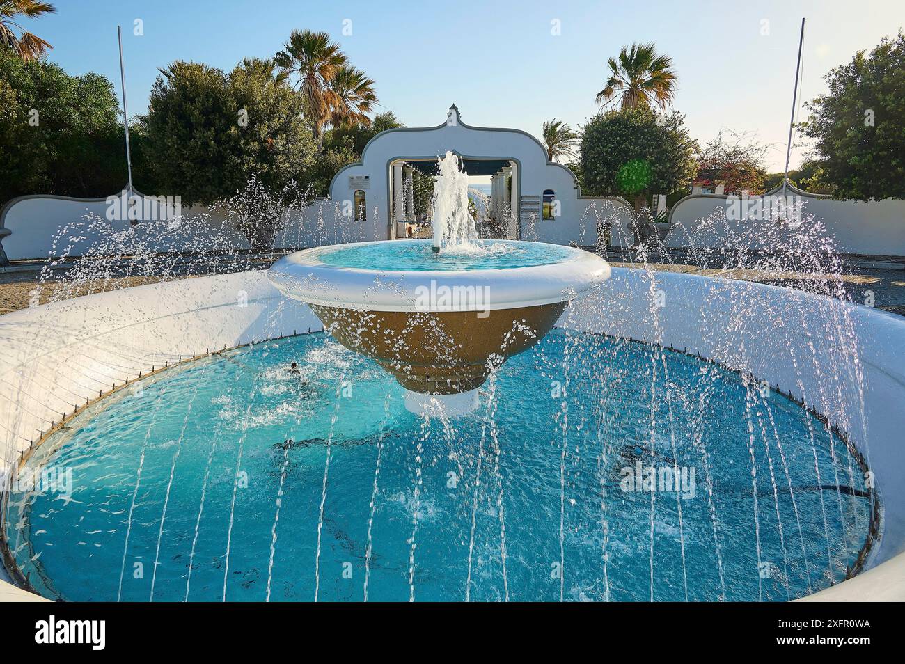 Symmetrical white fountain under palm trees, splashing water and sunny atmosphere, thermal ...