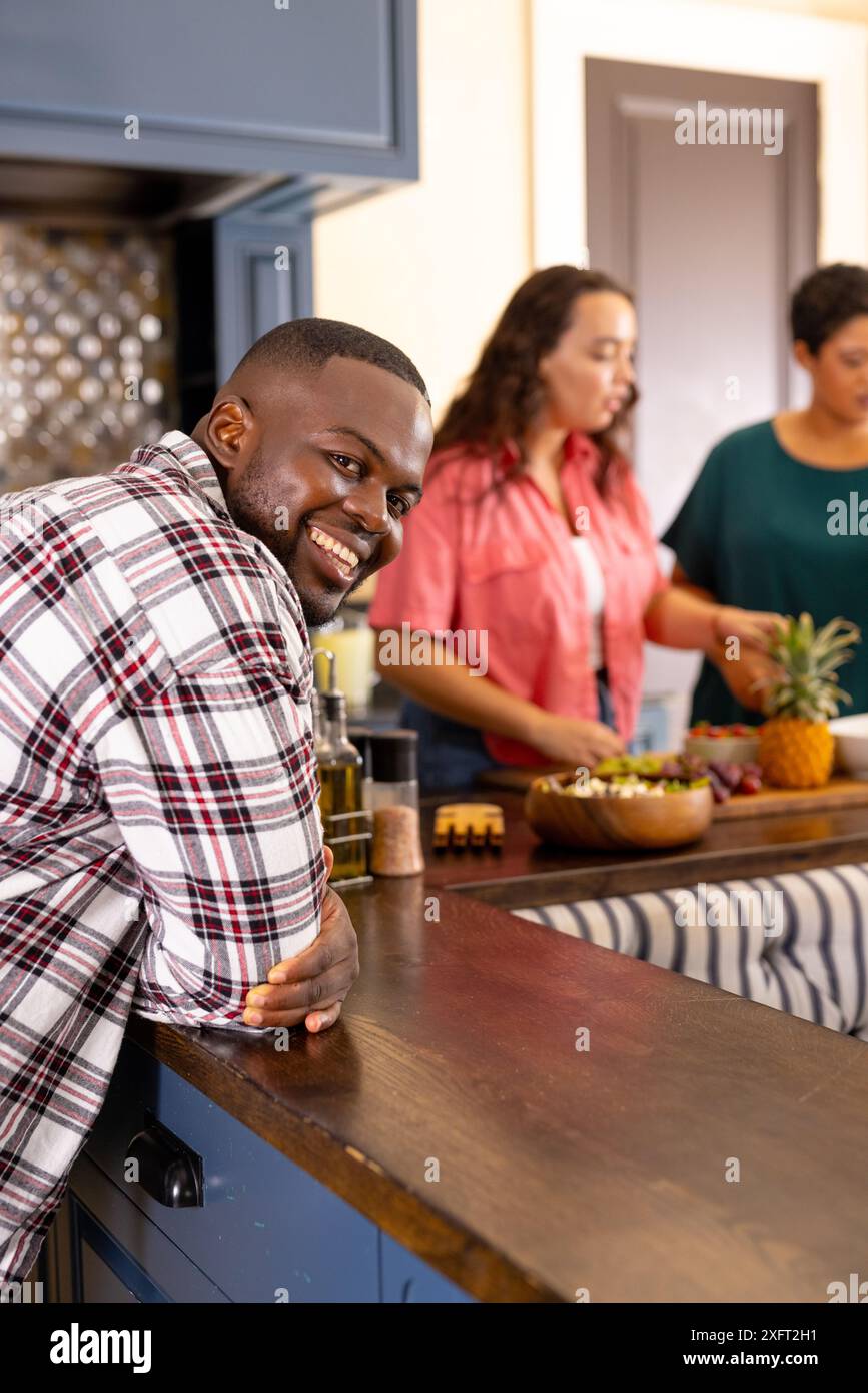 Smiling man leaning on kitchen counter while diverse friends preparing ...