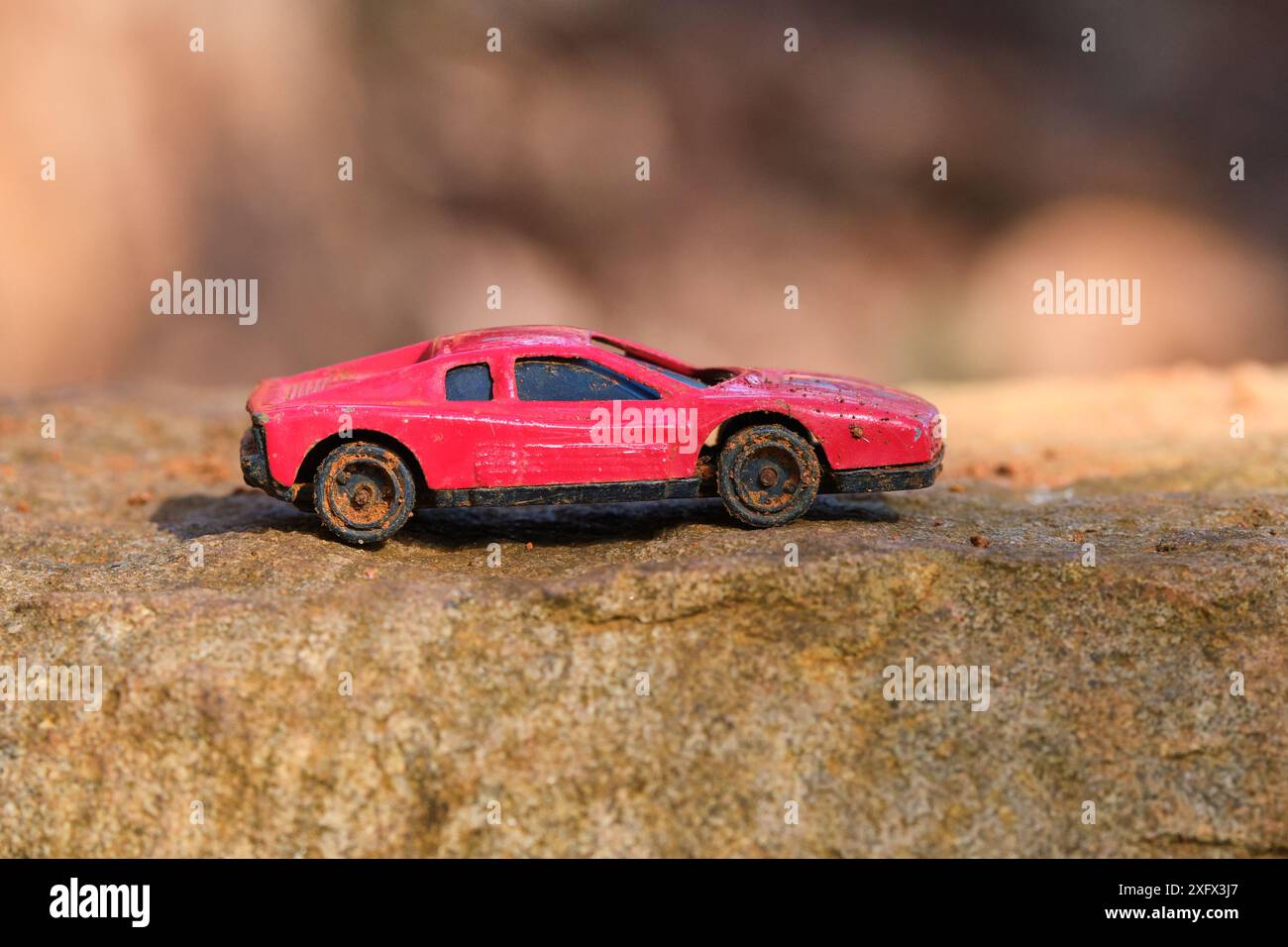 A close-up of a muddy, red toy sports car with damaged windows on rocky ...