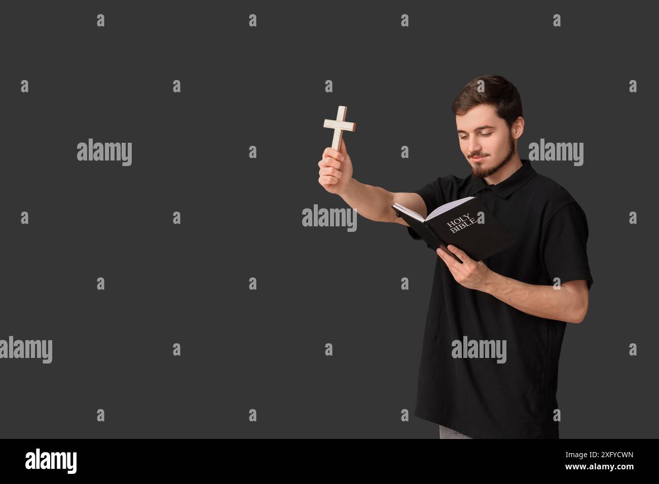 Religious young man with cross reading Holy Bible on black background ...