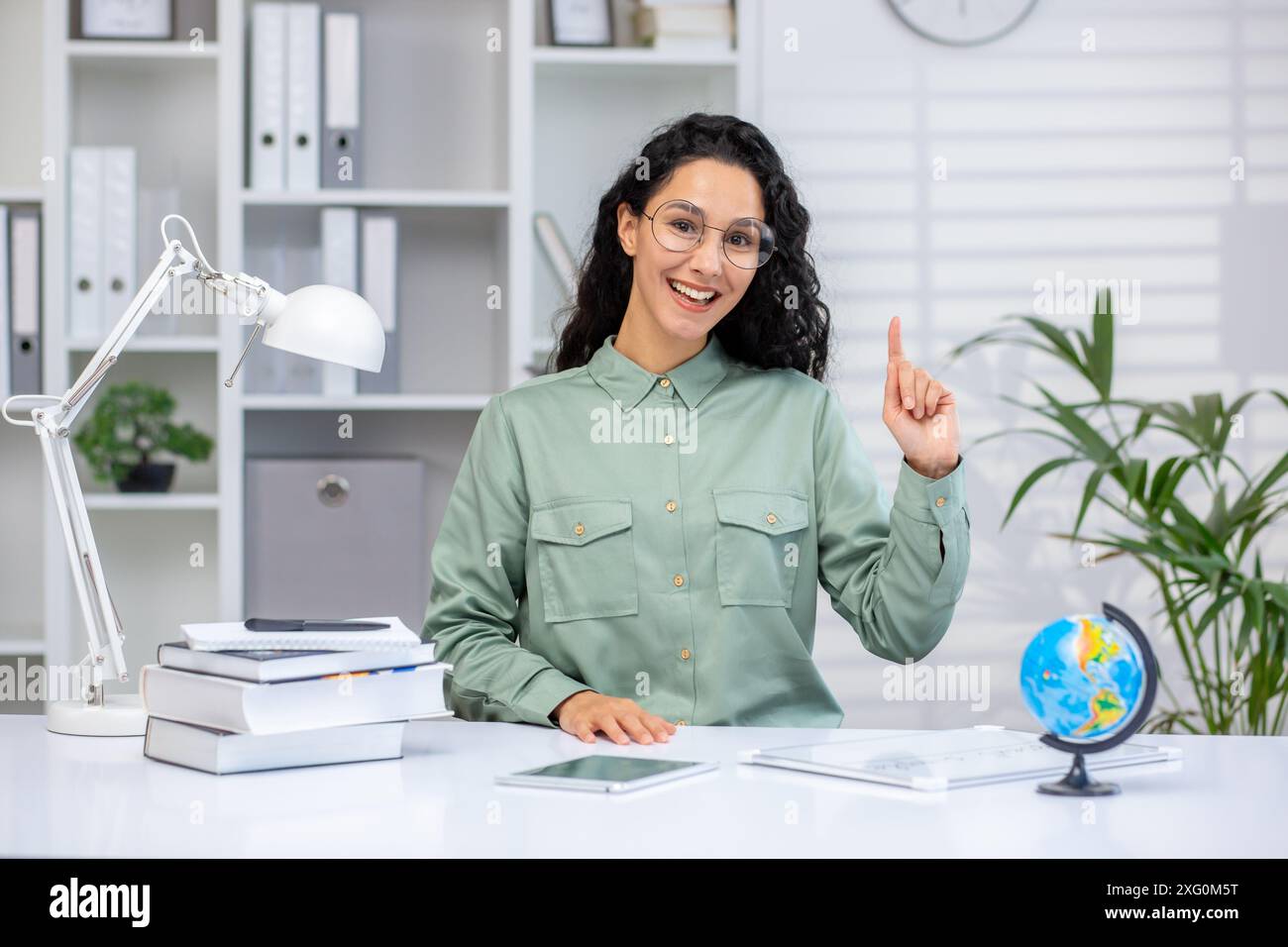 Cheerful female teacher raising her hand while sitting at a desk in a ...