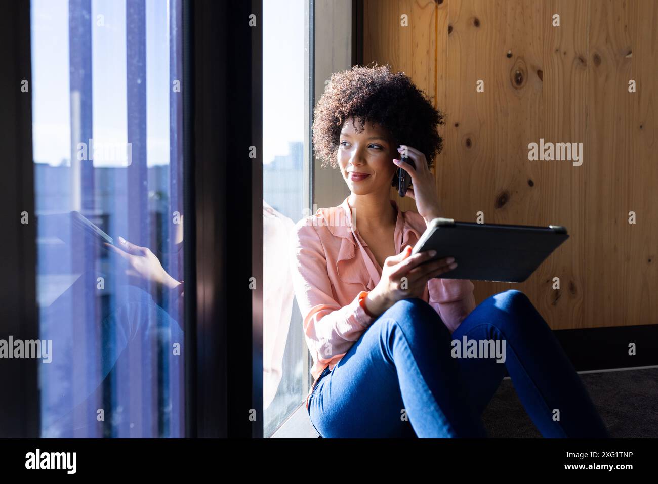 Talking on smartphone while holding tablet, woman sitting by window ...