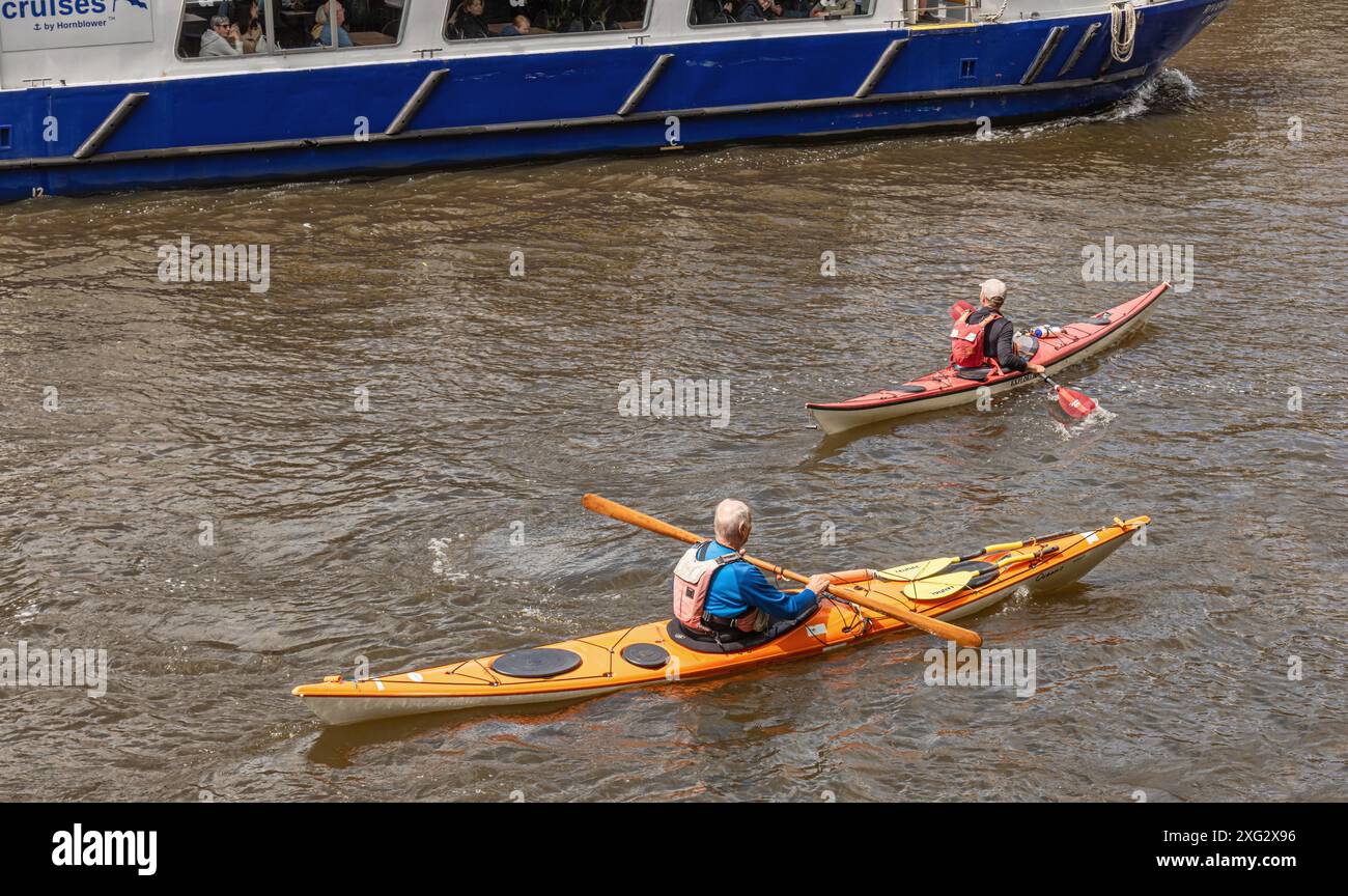 Two canoes paddle along a river together. In the background a tour boat ...