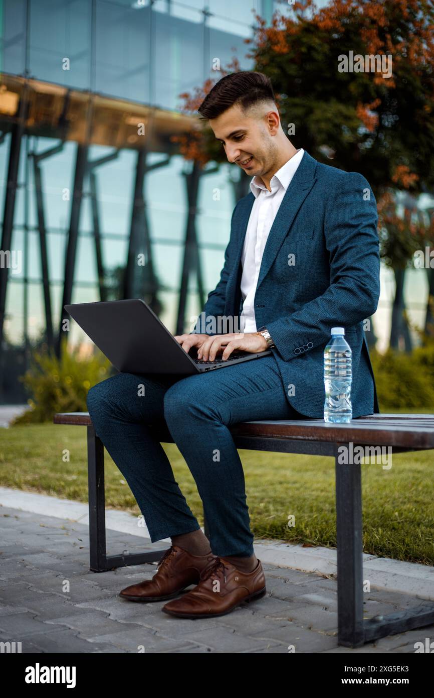 A professional man in a formal suit is seated on a bench while typing ...