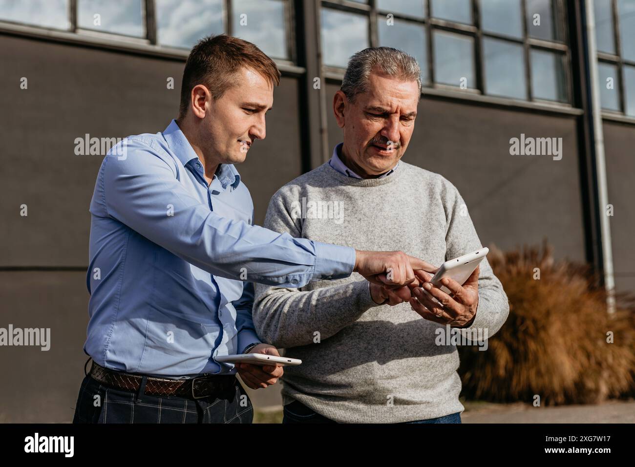 Two men stand outside of a building, one pointing at a tablet screen ...