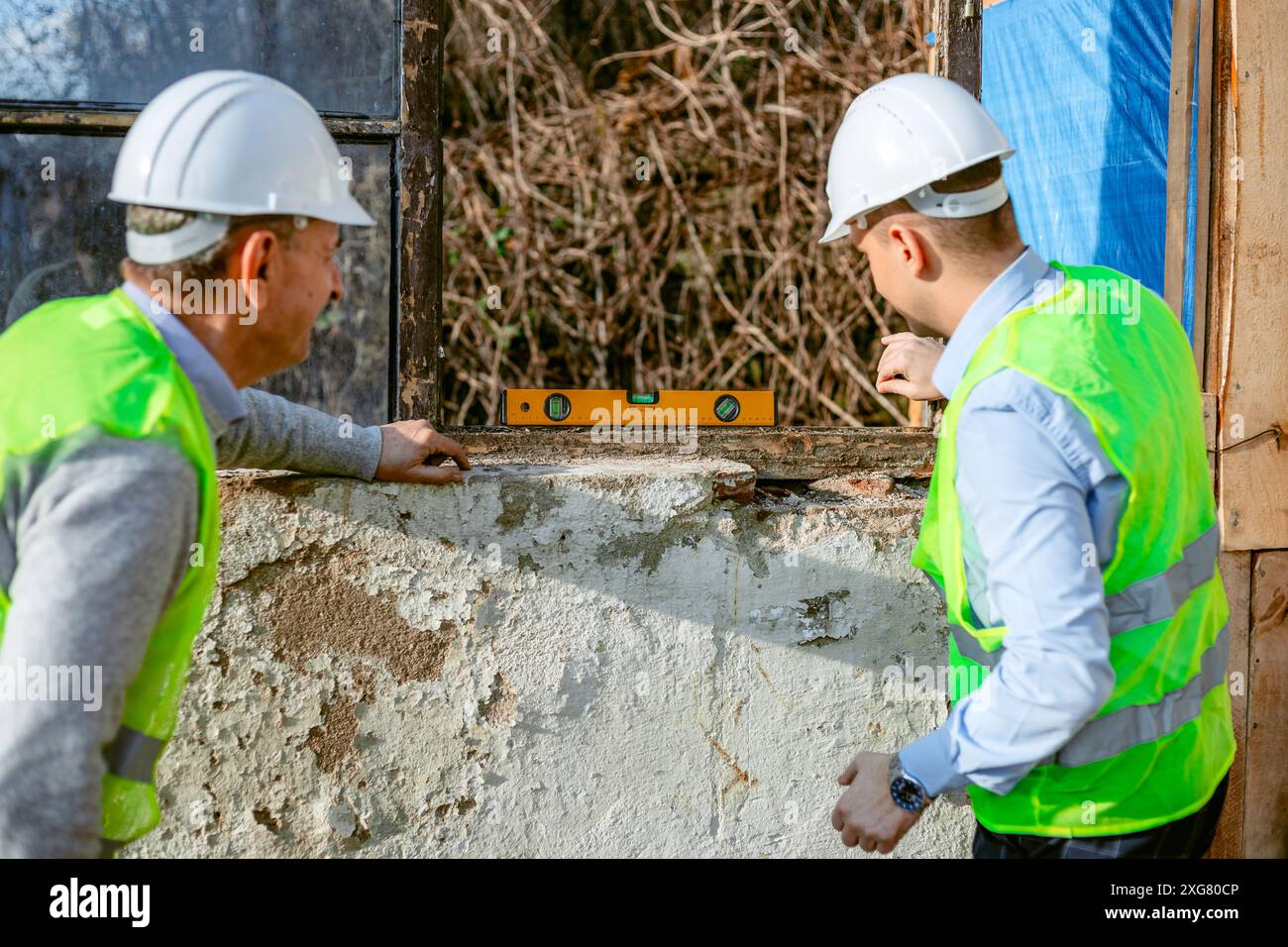 Two construction workers, wearing hard hats and safety vests, carefully check the level of a ...