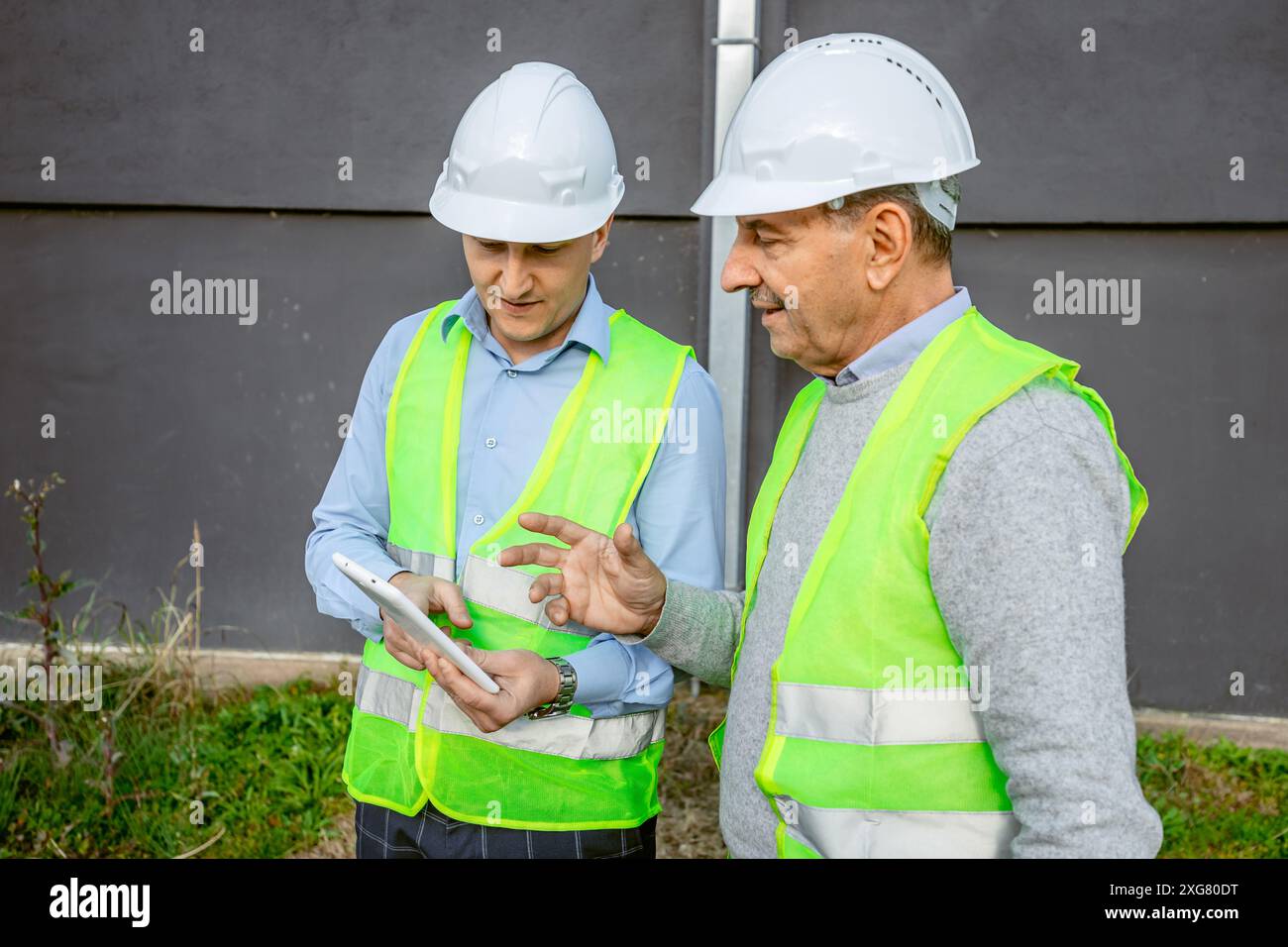Two construction workers wearing hard hats and safety vests stand outside a building during the ...