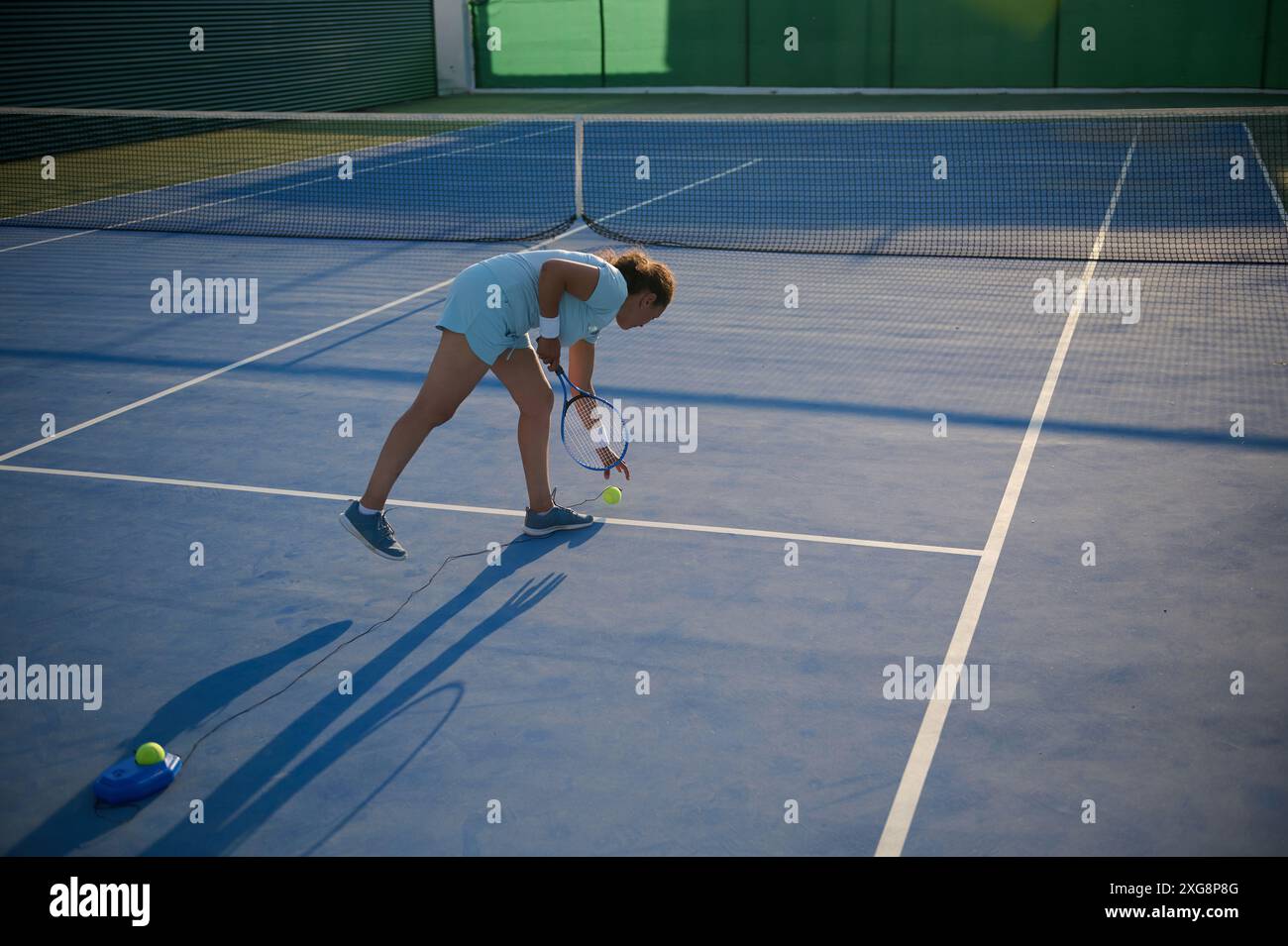 A young woman is practicing tennis on an outdoor court, bending down to ...