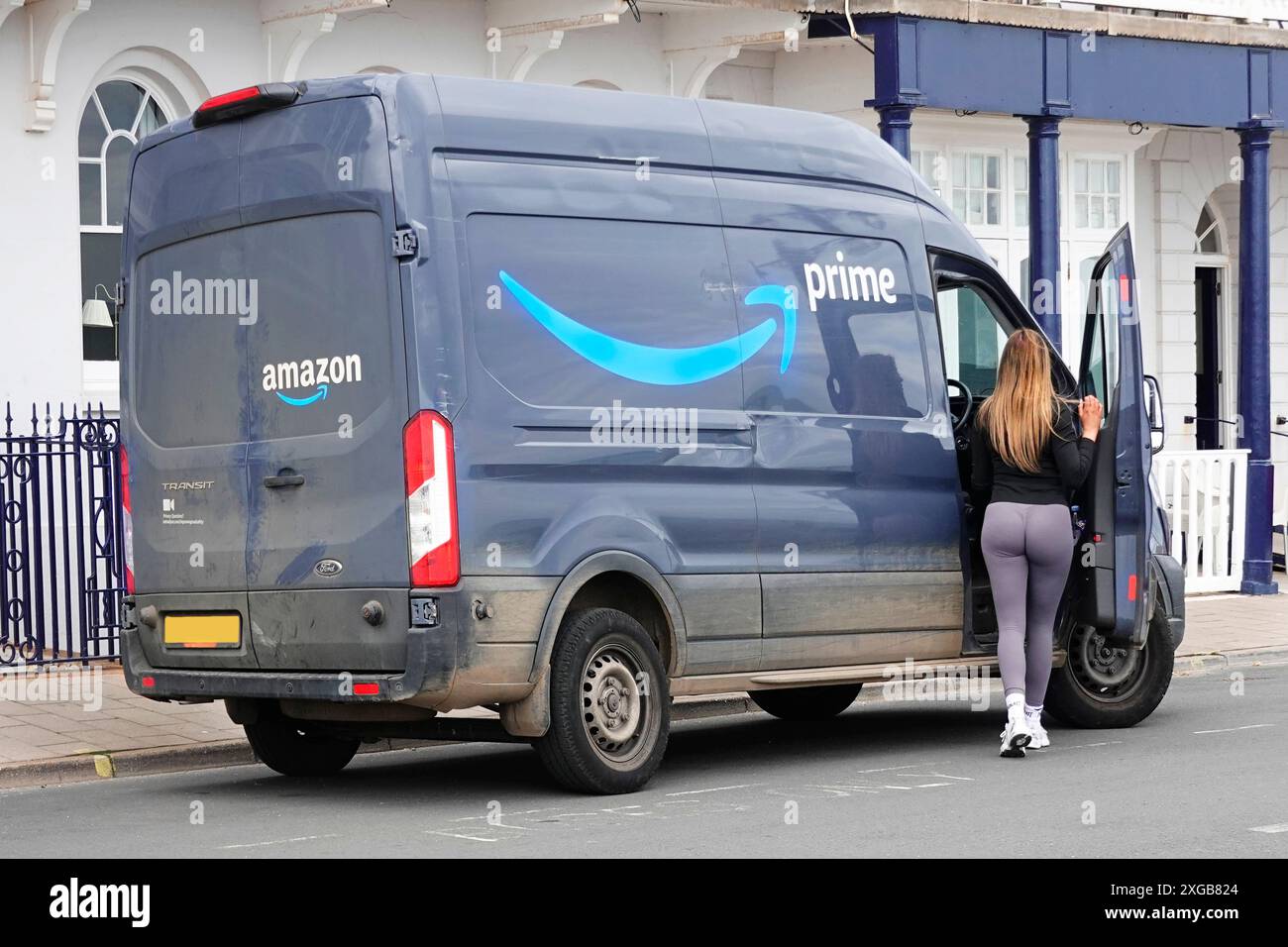 Woman Prime delivery driver back view returning to parked Amazon Ford van completing parcel ...