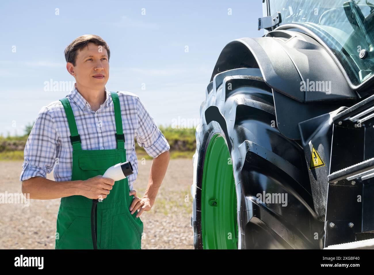 Farmer in green uniform stands next to electric tractor and holds ...