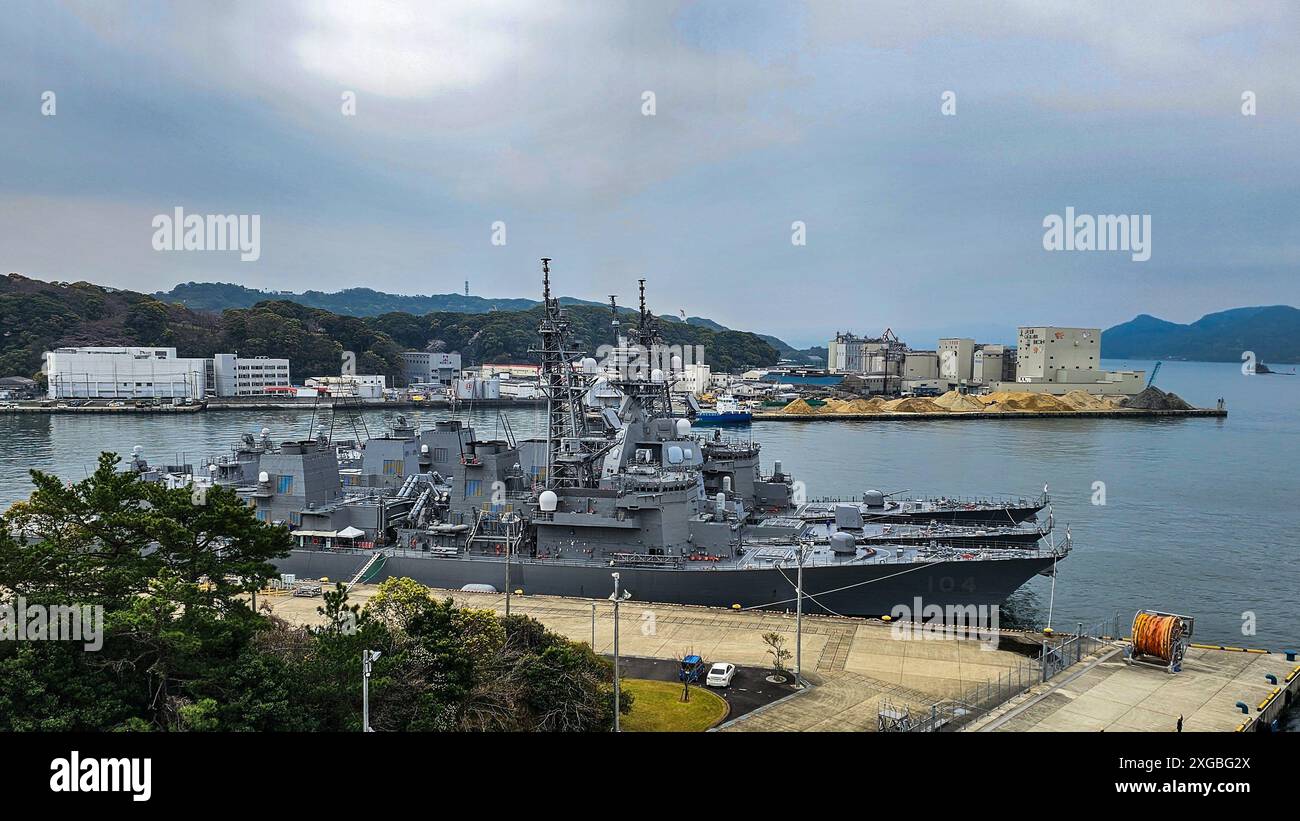 A naval ship docked at a harbor with industrial buildings and hills in ...