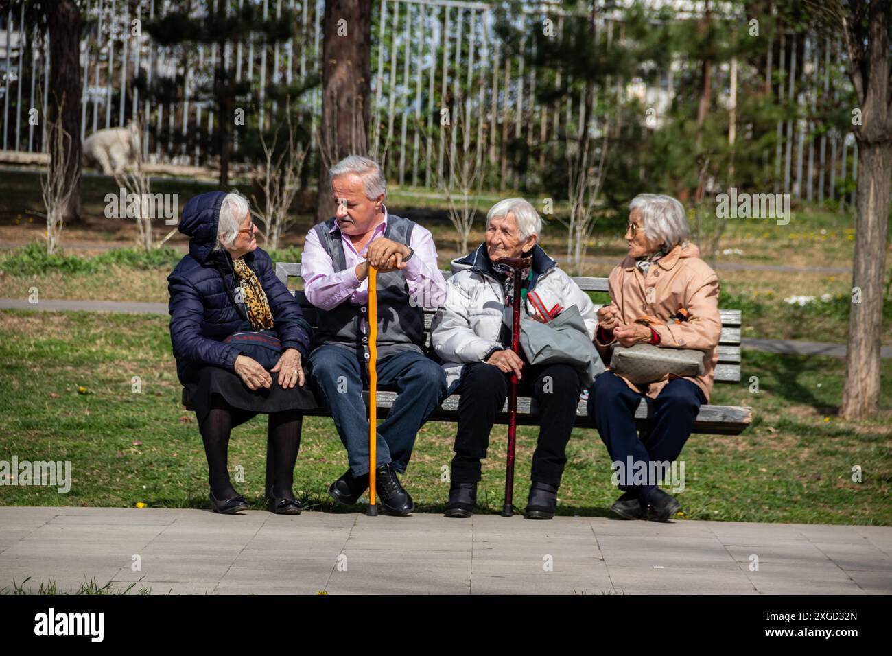 Several retired people sit on a bench in a public city park, enjoying the sunny weather ...