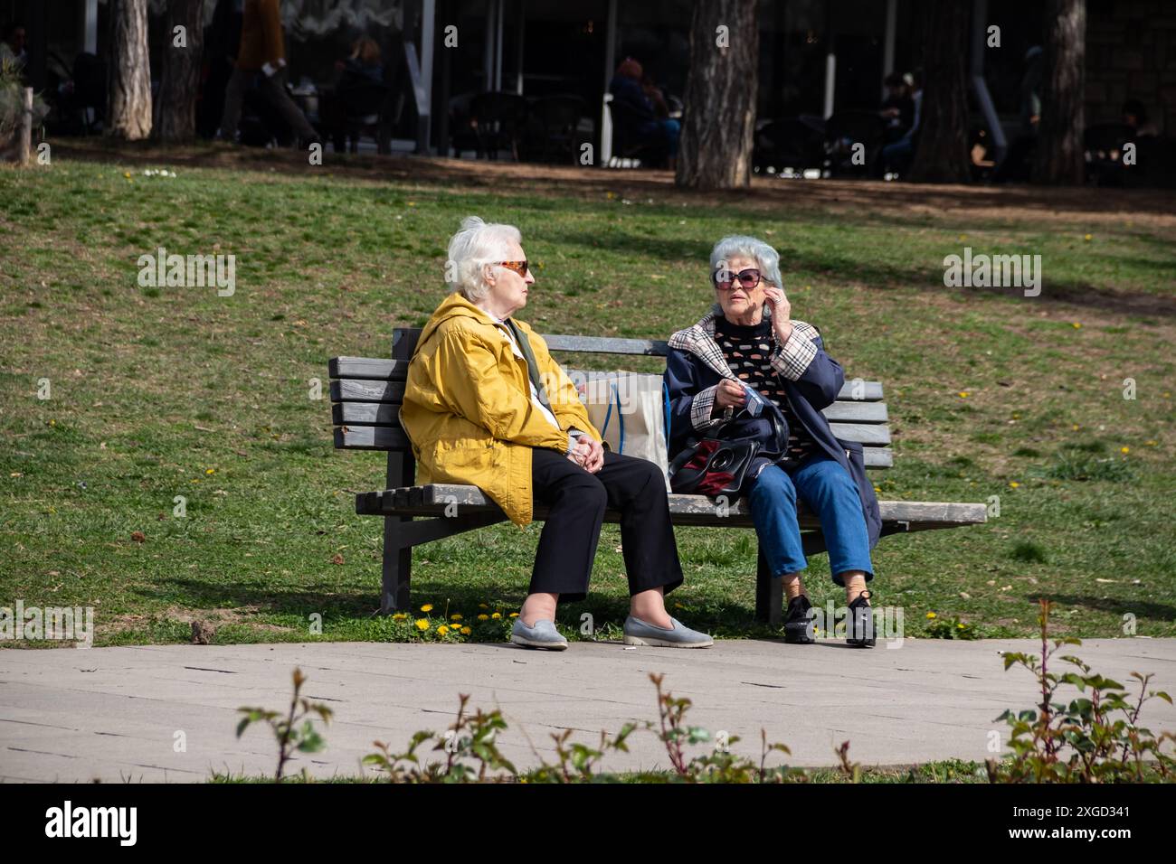 Several retired people sit on a bench in a public city park, enjoying the sunny weather ...