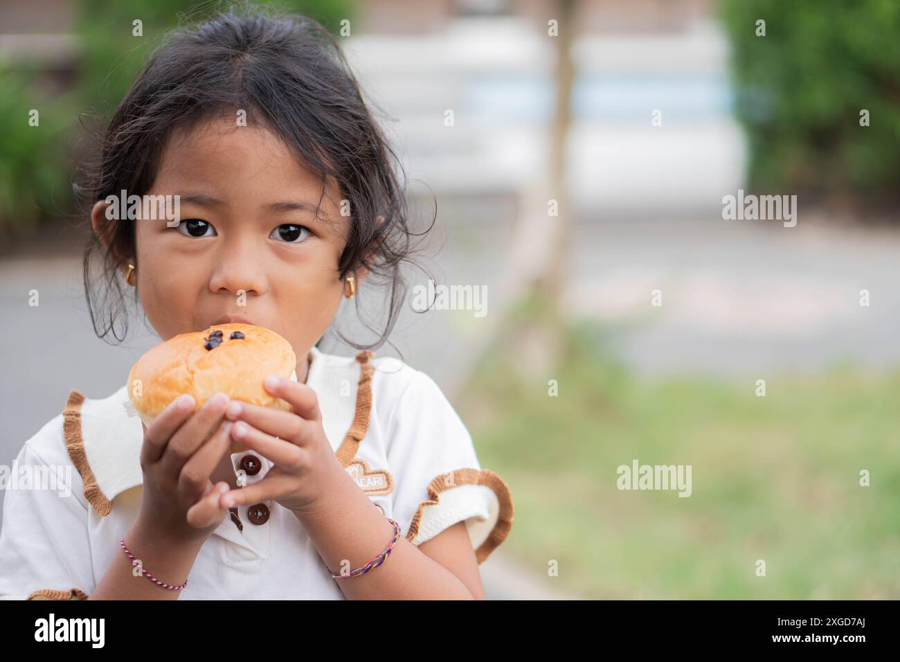A young girl eating a bun outdoors, with a blurred background of ...