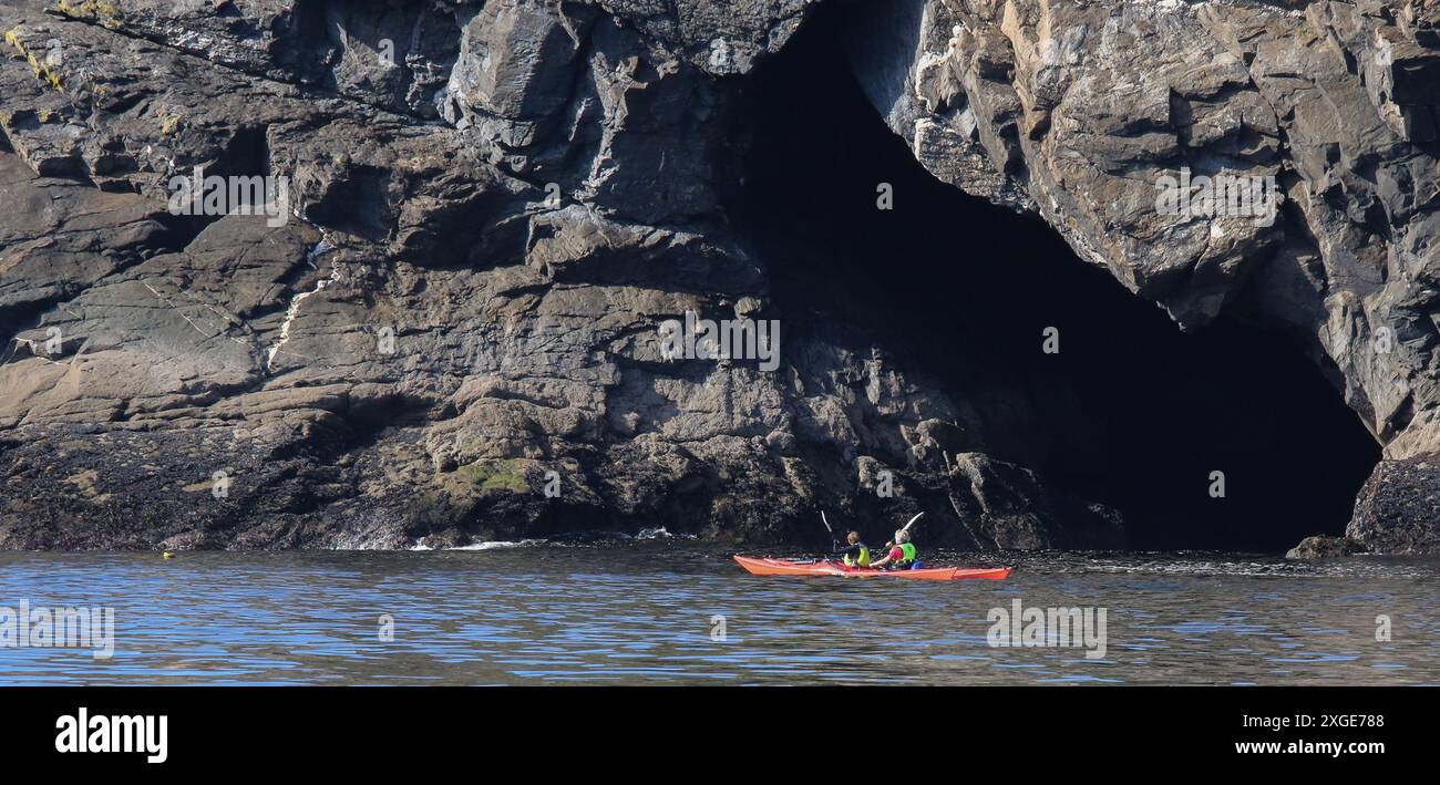 Two kayakers paddling red kayaks past mouth of sea cave coast of ...