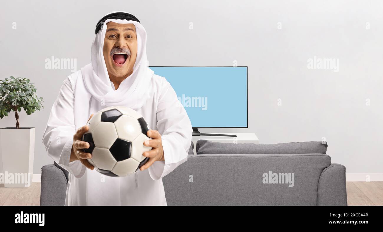 Excited muslim man holding footballball in a room with tv and sofa ...